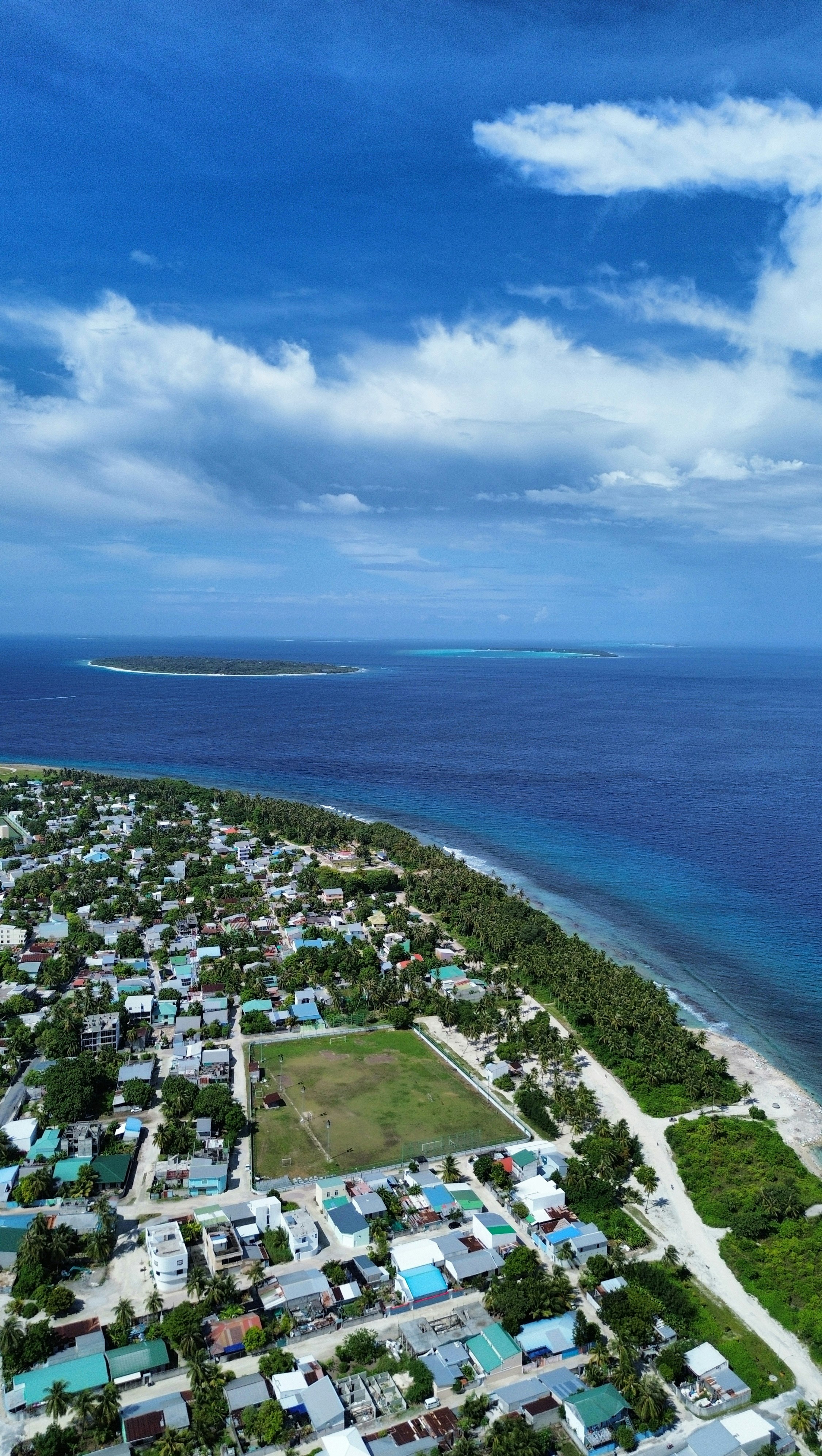 Aerial view over a tropical coastal town with a long shoreline and turquoise waters, showing a grid of colorful rooftops and a central grassy field under a bright sky.