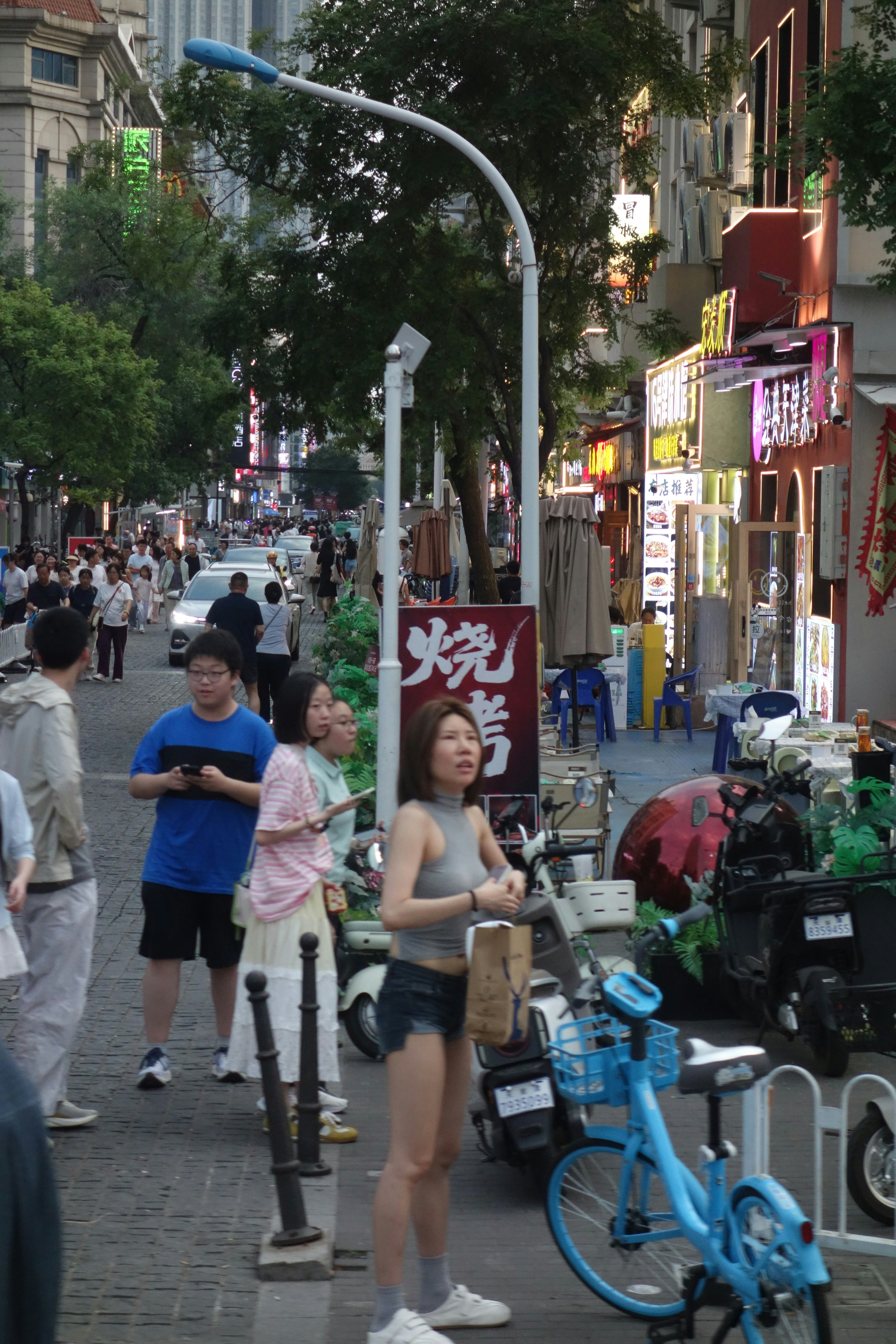 Bustling urban street scene with a young woman in the foreground and neon-lit storefronts along a pedestrian boulevard at dusk.