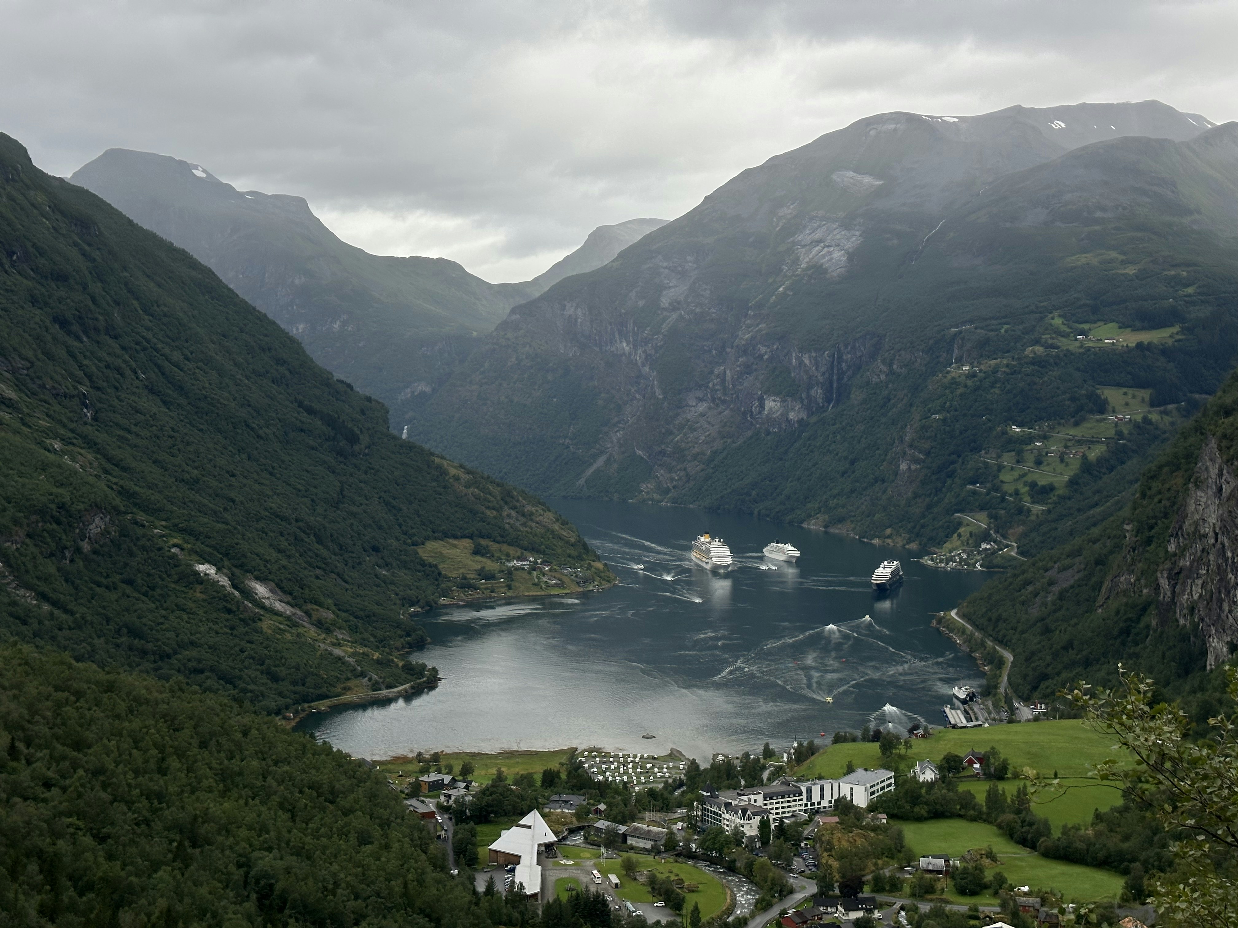A valley village sits along a calm fjord with steep green mountains rising on both sides and a few icebergs floating in the water.