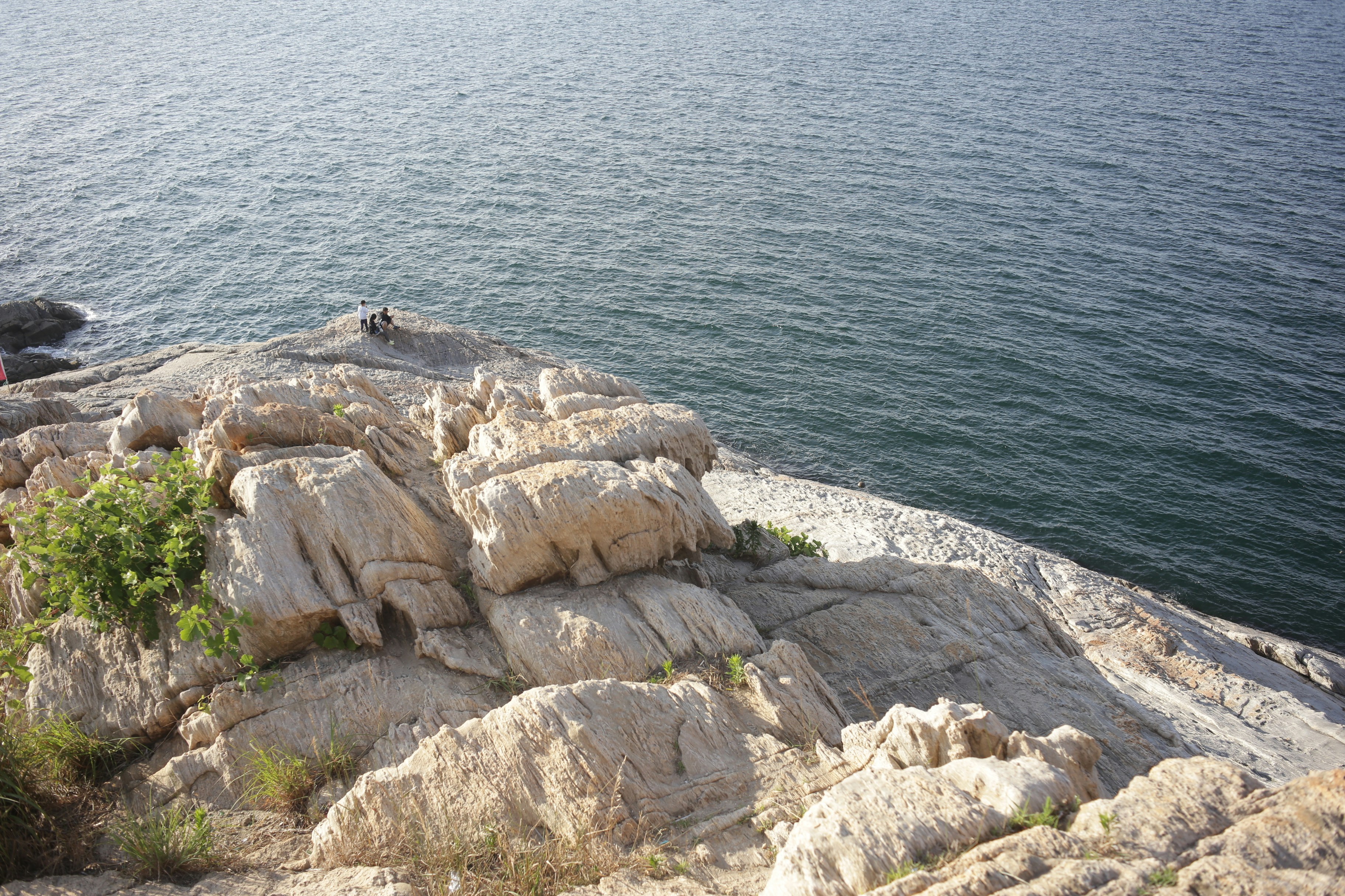 Rugged coastline rendezvous: two hikers perched on sunlit limestone ledges above a quiet sea