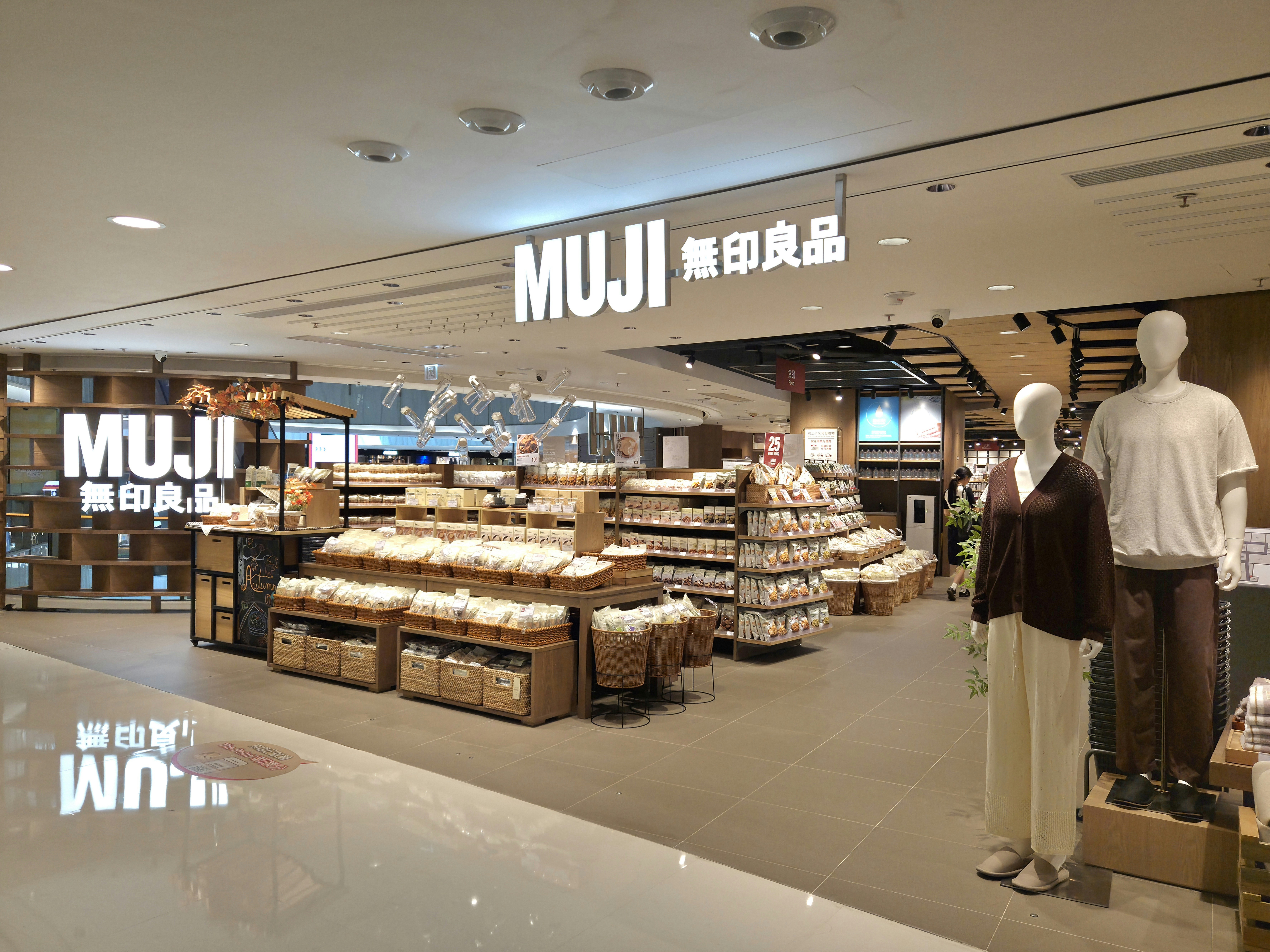 MUJI storefront within a shopping mall, featuring neatly arranged product displays and two mannequins near the entrance in soft ambient lighting.