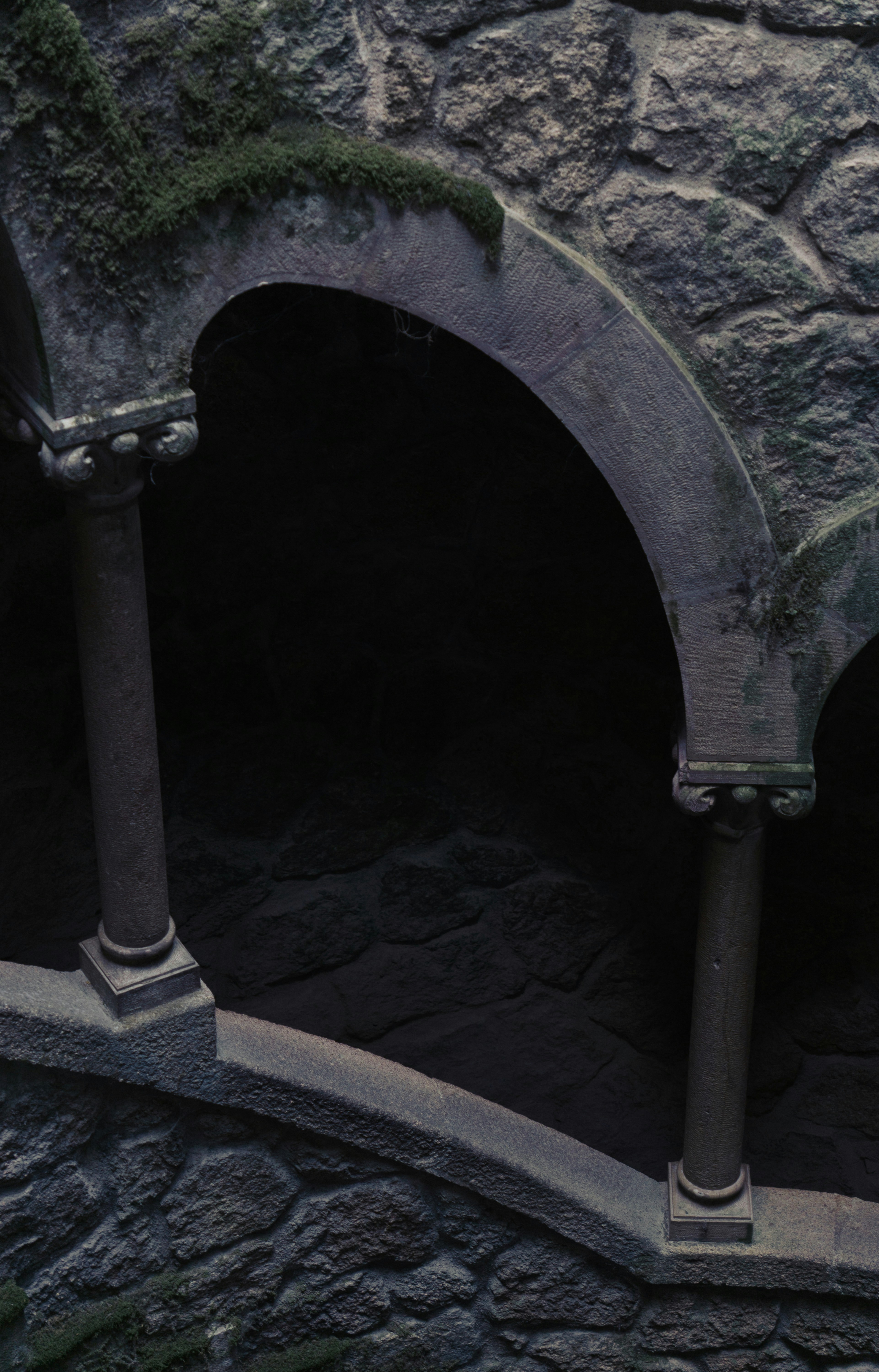 Dimly lit stone arch with moss-covered top and vertical iron columns, emphasizing architectural texture and aged materials.