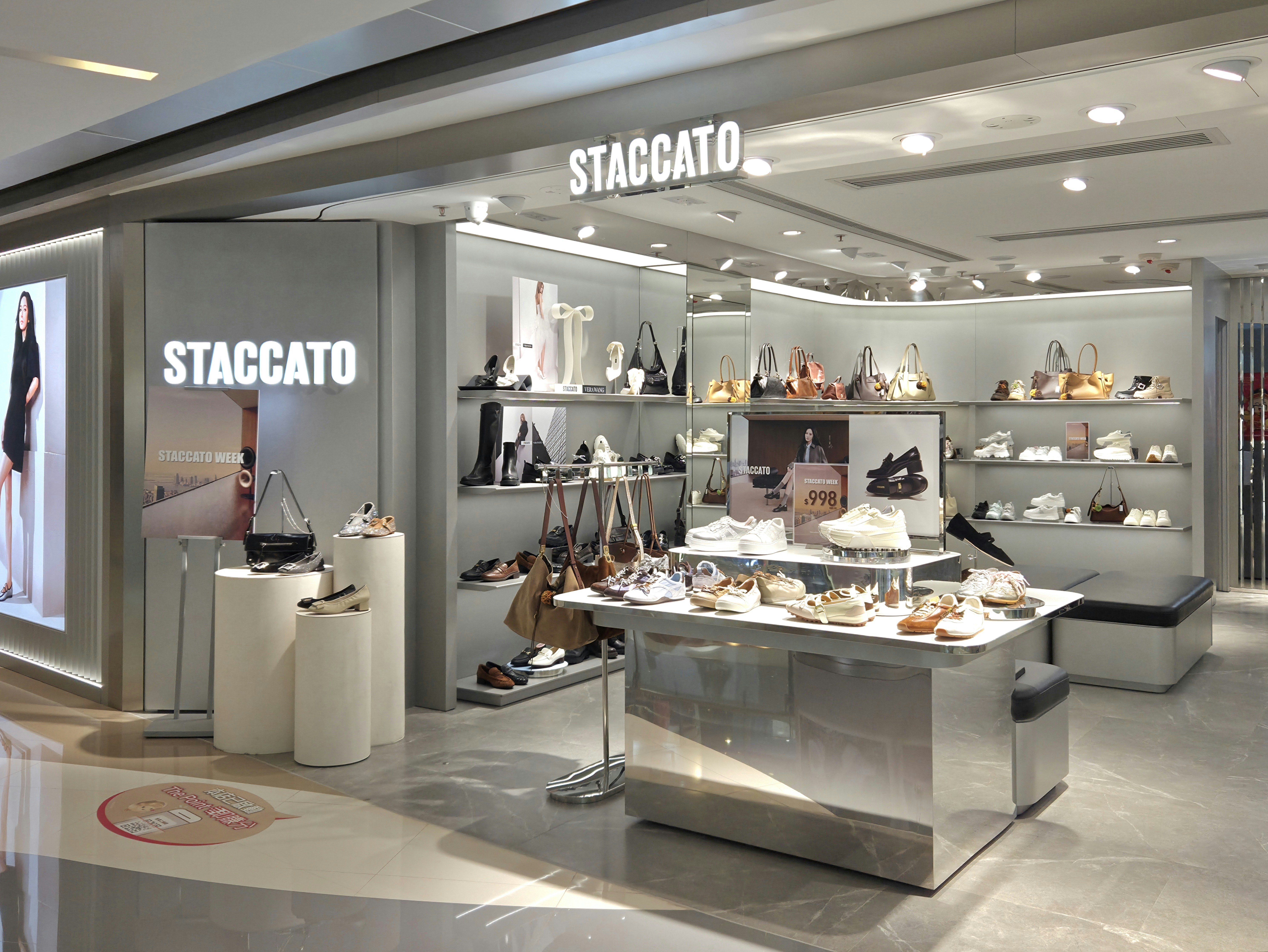 Shoe store display with bright signage and organized shelves inside a mall corridor. A central glass table showcases various styles while a bench sits nearby, framed by a clean, well-lit storefront.