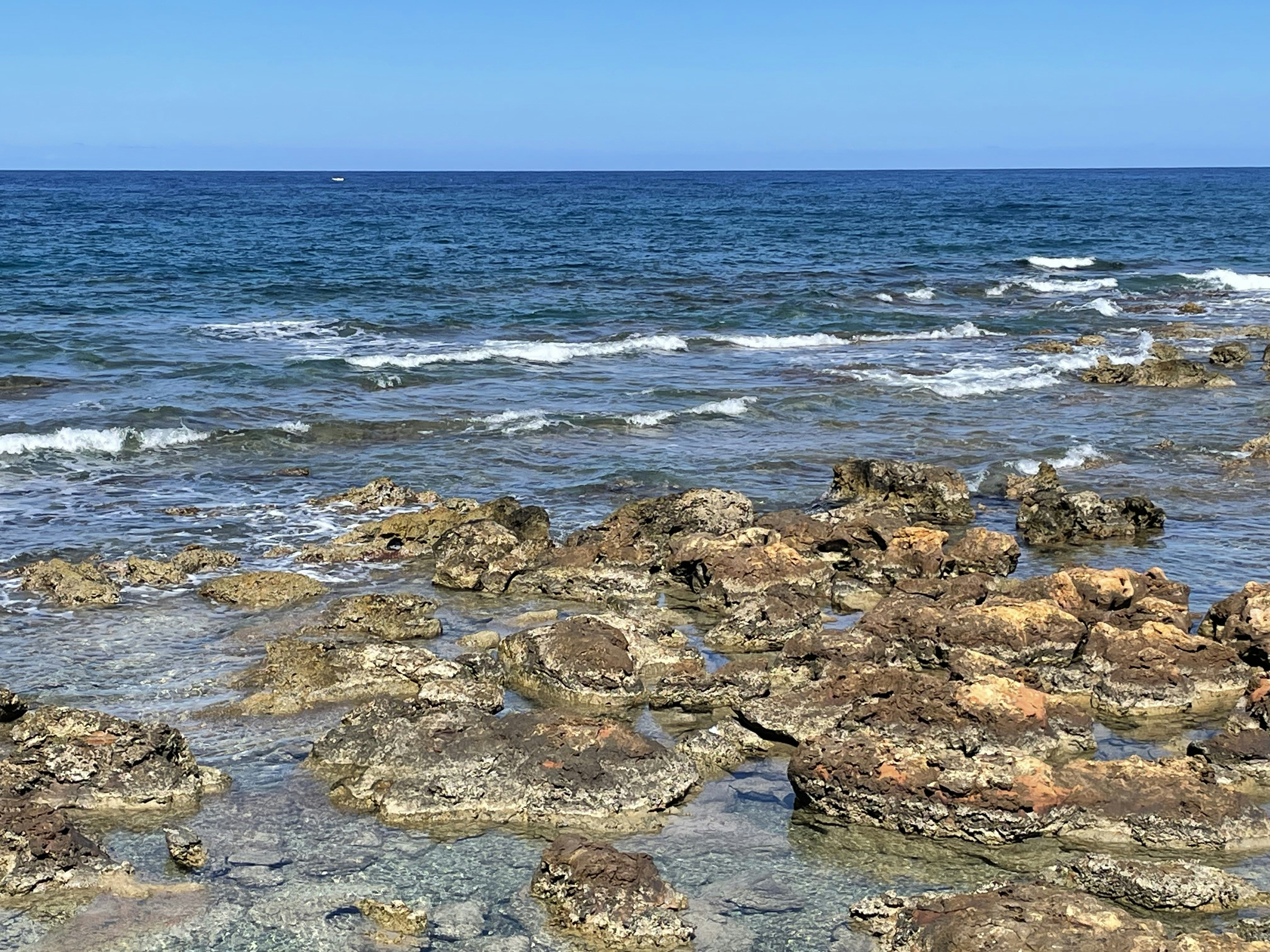 Sunlit rocky shoreline with turquoise water and gentle waves; a calm seascape emphasizing texture and contrast of rocks and sea.