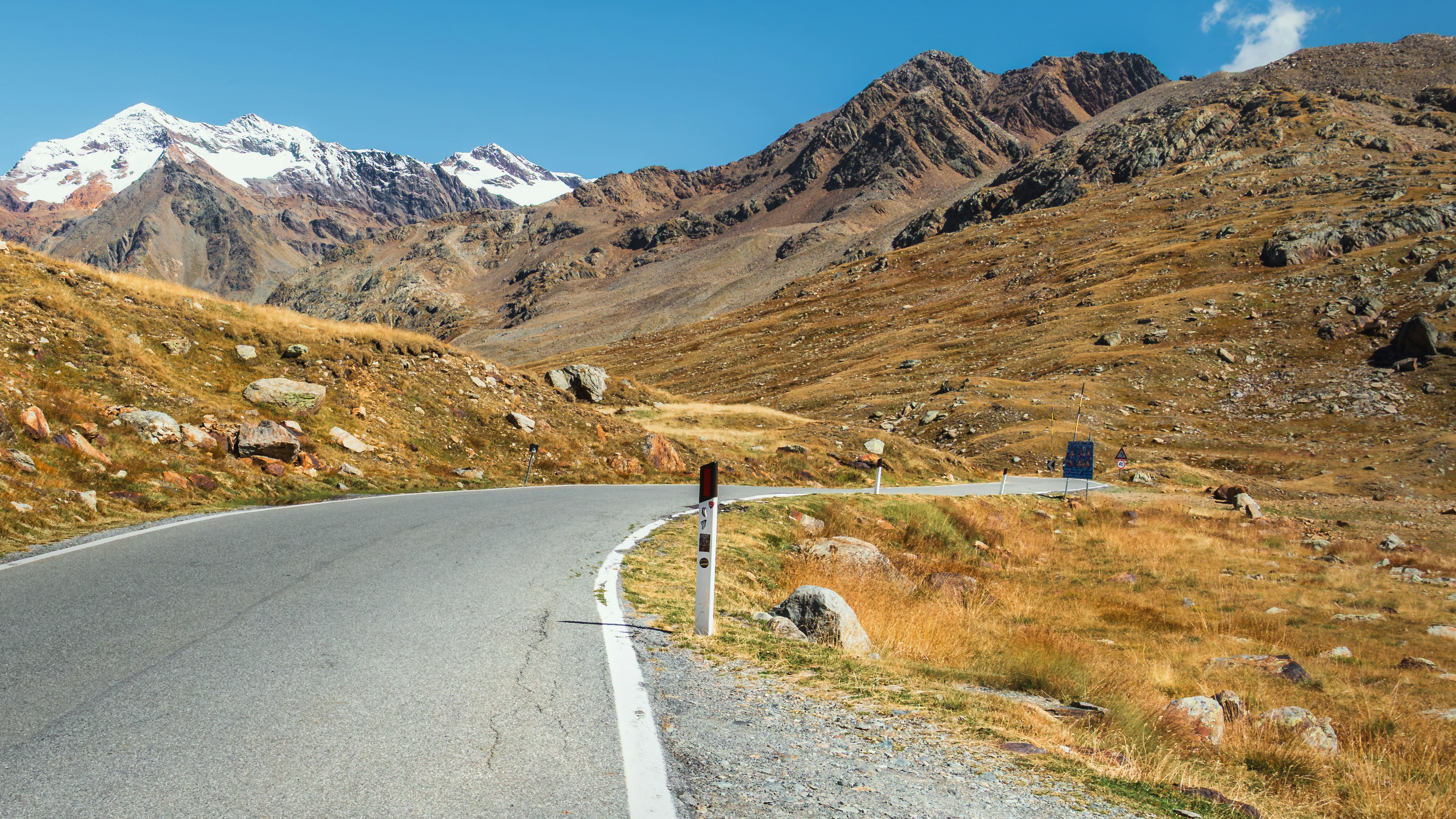 A winding mountain road cuts through barren alpine terrain with distant snow-capped peaks under a clear blue sky. The scene emphasizes rugged landscape and open travel.