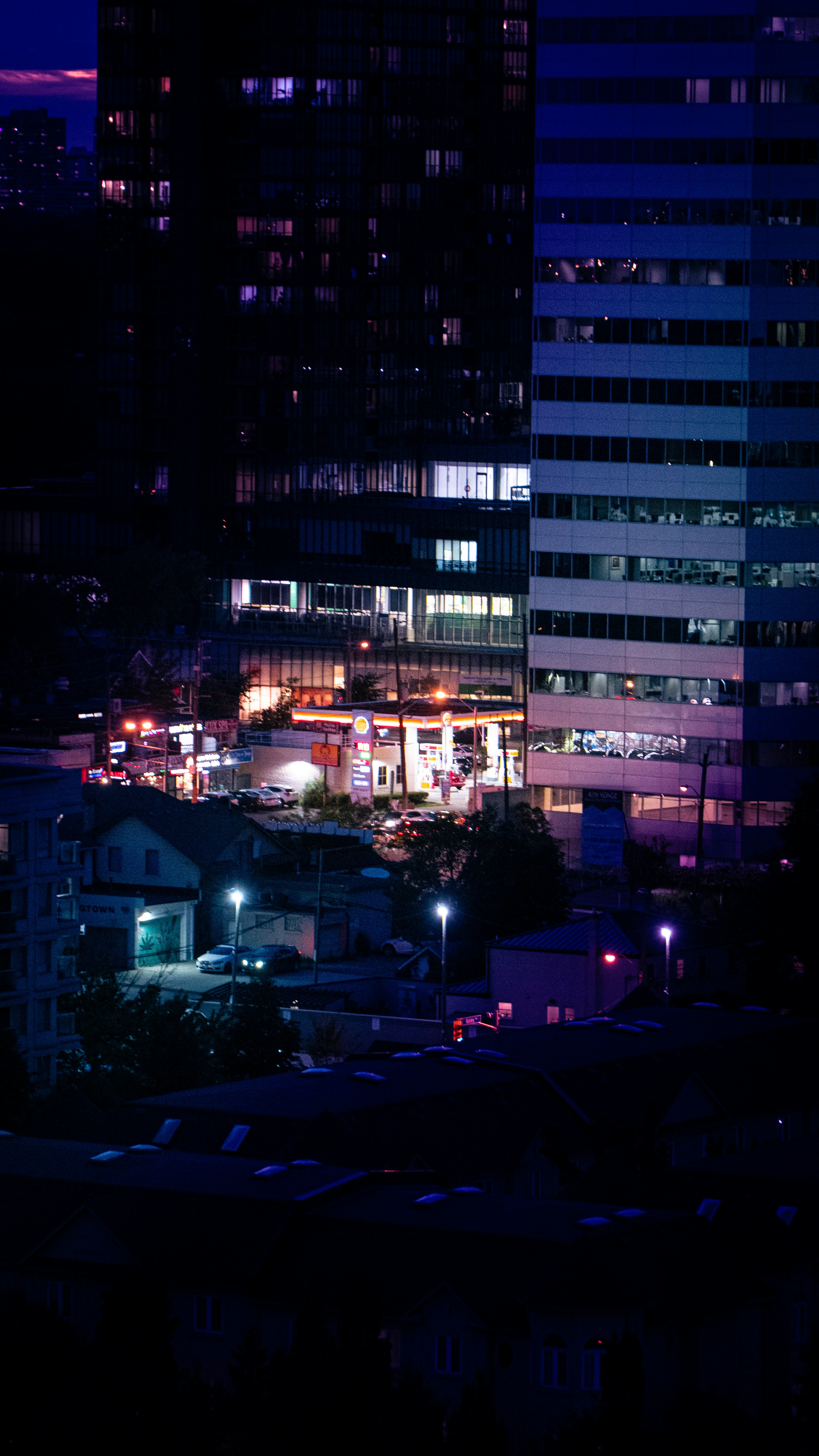 Tall city lights and a glowing gas station amid midrise buildings at night, with cool blue accents and warm signage tones.
