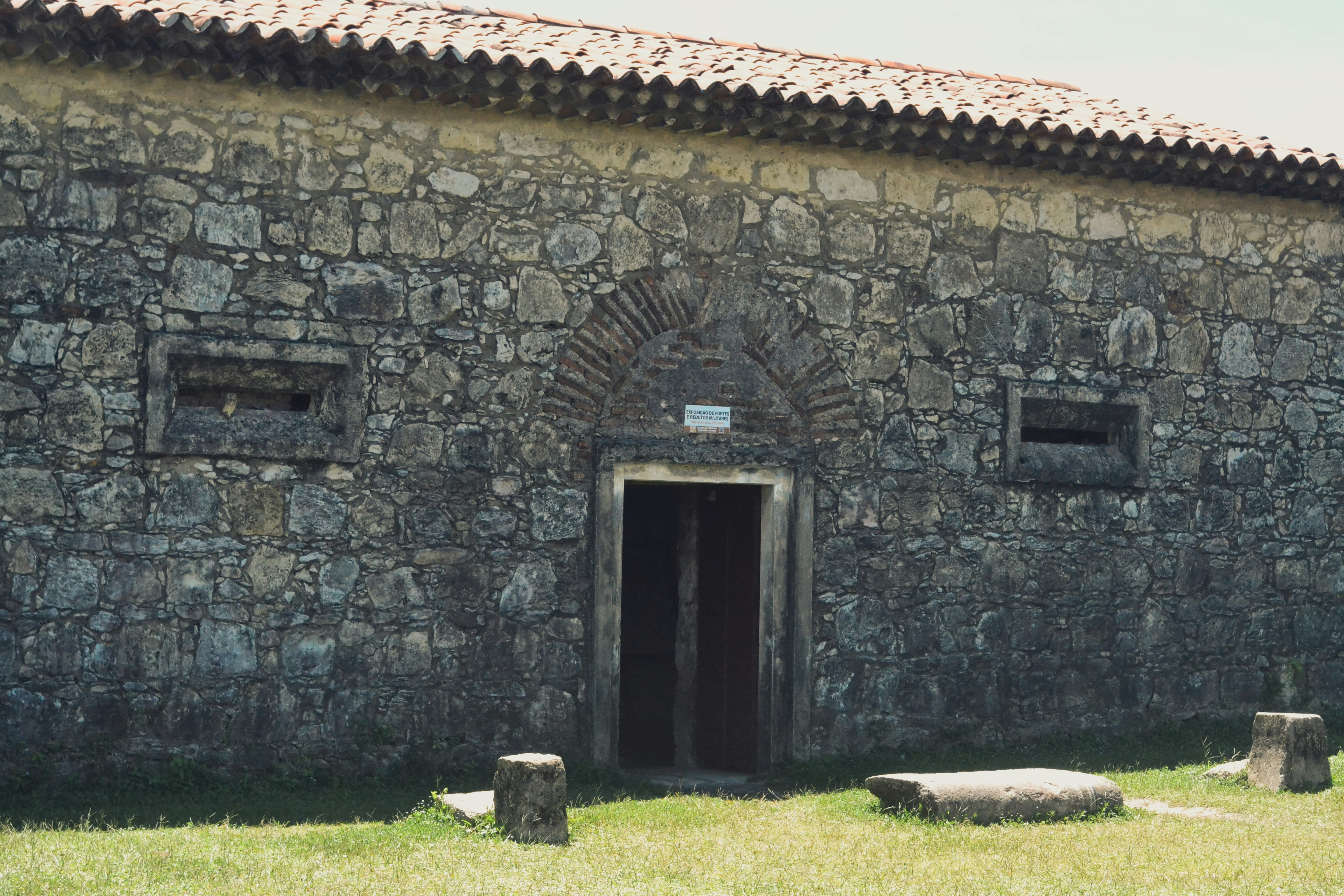 Weathered stone building with a central arched doorway and small side windows, set in bright grass under a tiled roof; an architectural relic revealing rugged textures and a muted, historic mood.