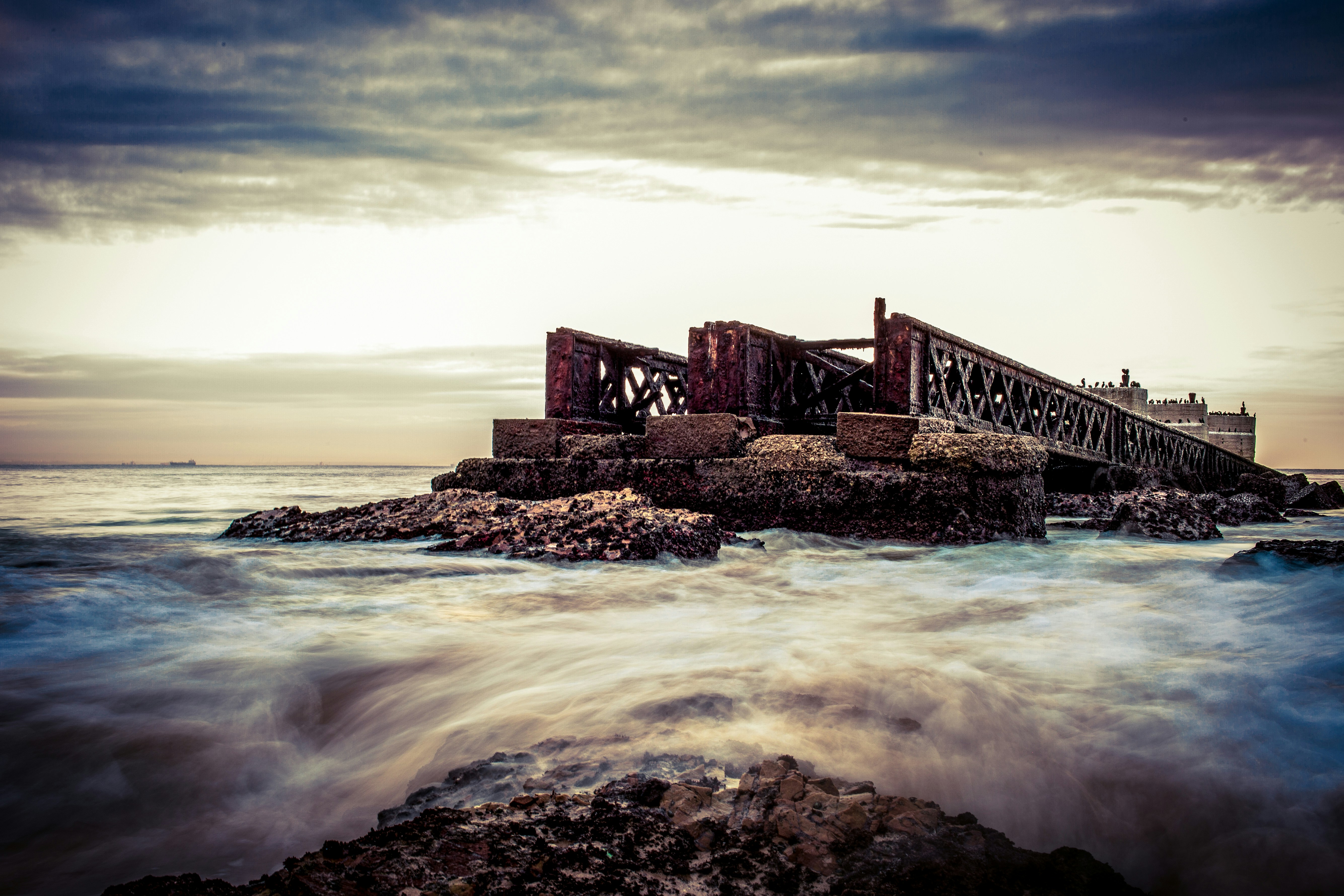 Weathered pier extends into calm sea under a moody sky; long exposure softens waves around rocky foreground.