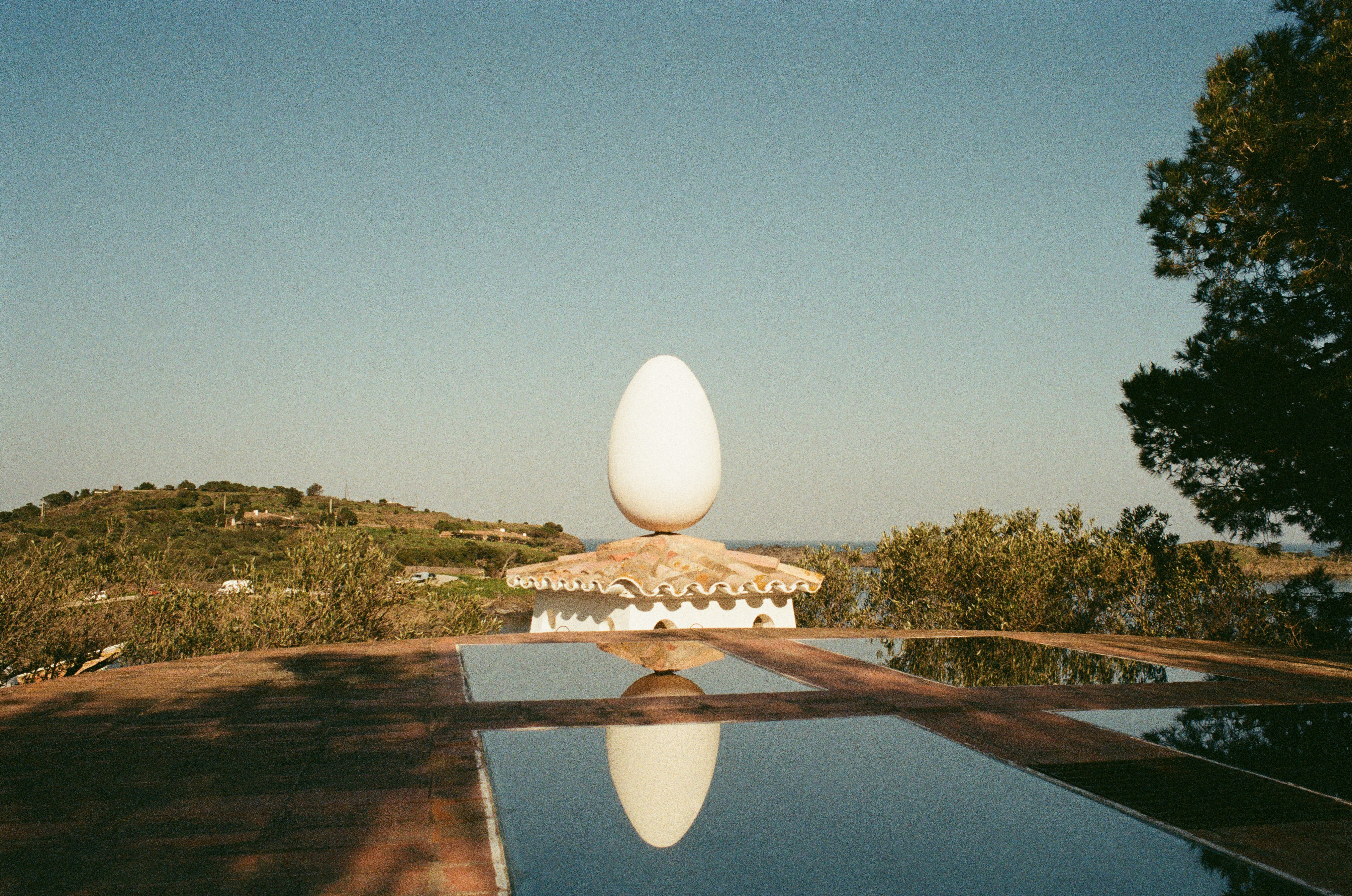 A smooth egg sculpture sits atop a decorative pedestal reflected in a narrow pool on a sunlit terrace, with distant hills and a tree on the right. A calm, serene outdoor scene focused on symmetry and stillness.