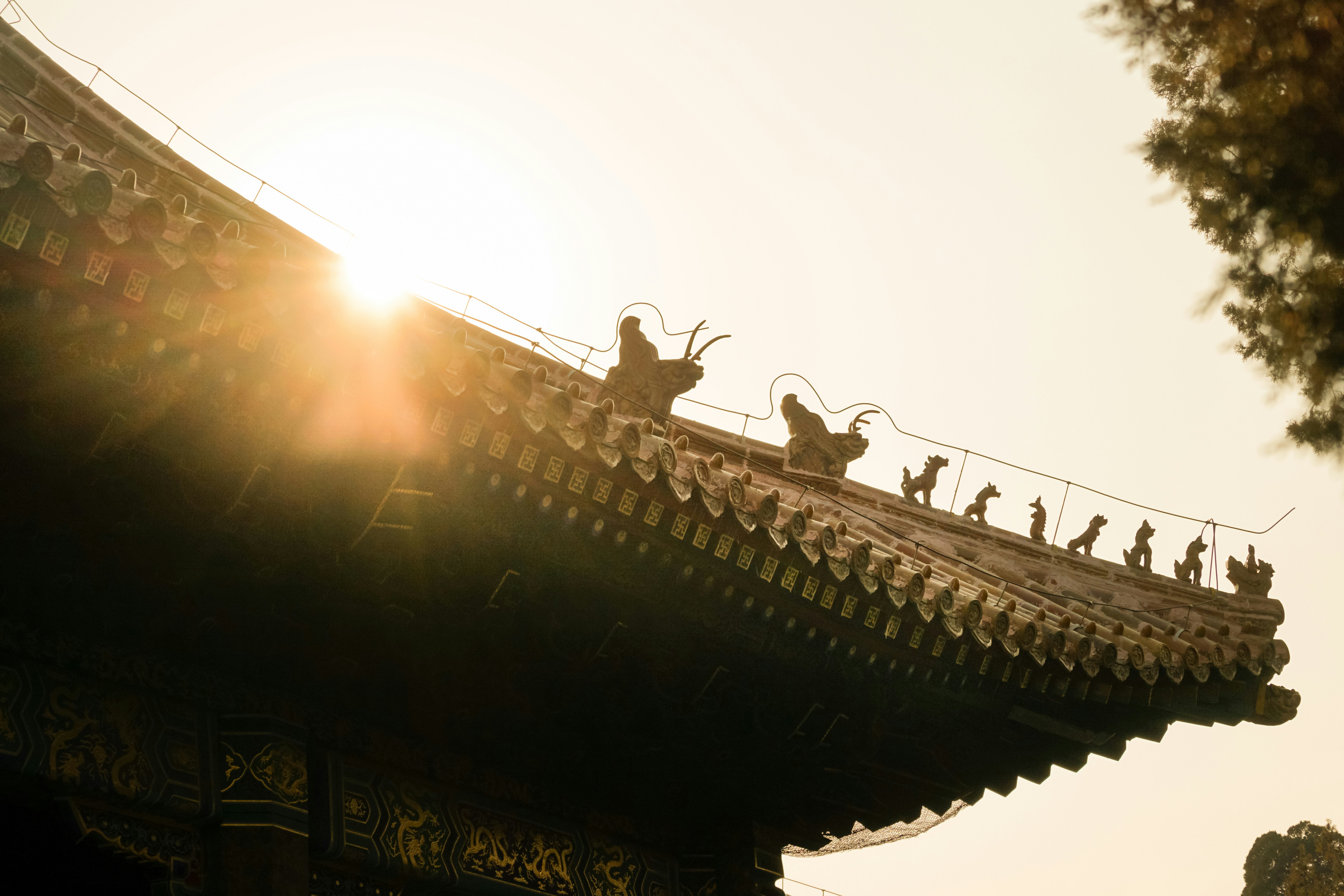 Sunlight streams over a traditional tiled roof, silhouette figures line the edge as decorative guardians stand against a pale sky.