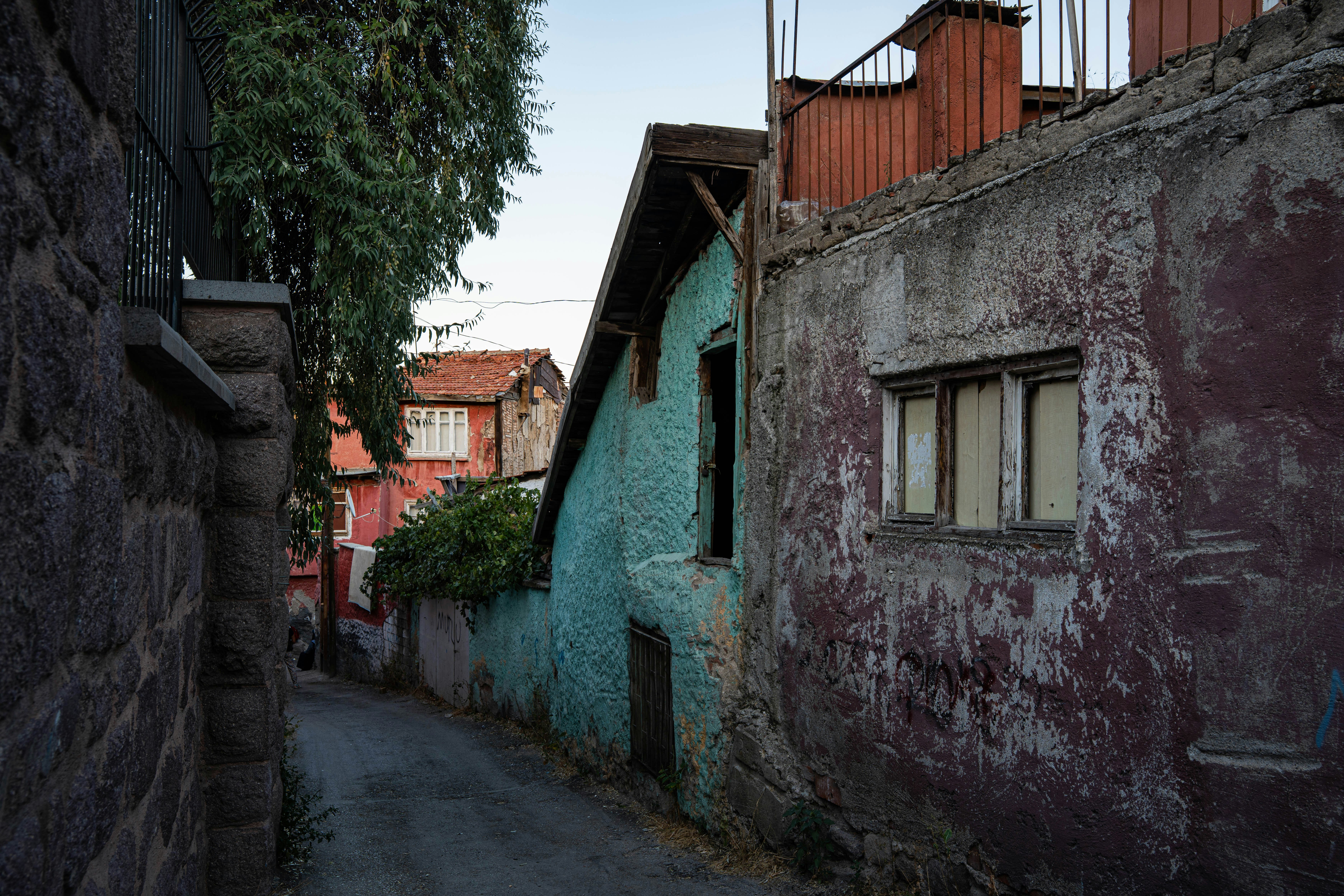 Vividly colored walls line a narrow alley, with a calm, shadowed street inviting the viewer to explore deeper shadows and distant light. A mural-like turquoise house stands out among weathered facades in a quiet urban lane.