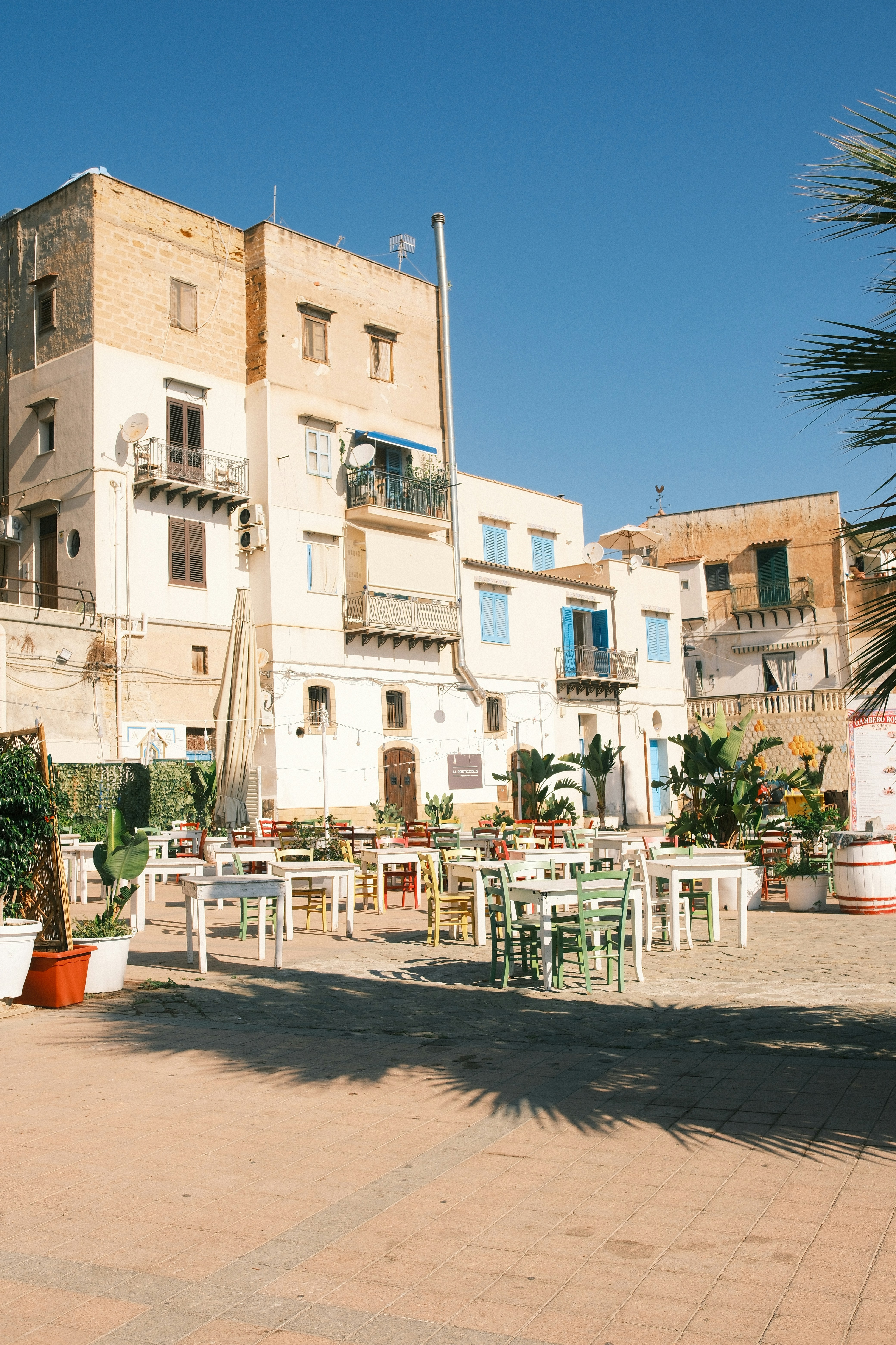 Sun-washed outdoor dining area with colorful chairs and tables lining a sandy plaza in front of a row of pastel buildings against a clear blue sky. Note: the scene includes a cafe area with plants and shadows from palm fronds.