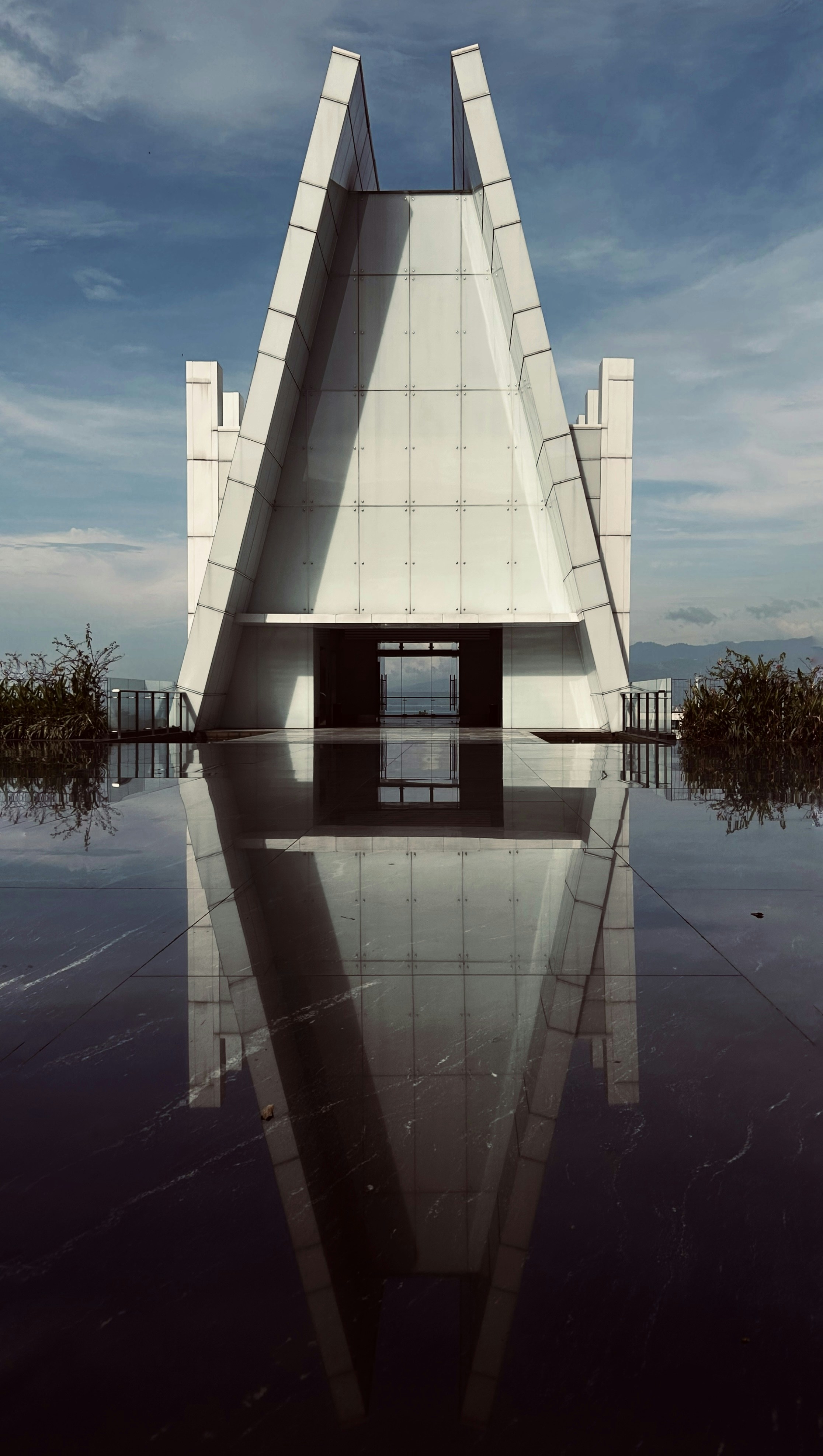 Architectural structure with dramatic inverted V shapes rising above a reflective pool, creating a crisp vertical symmetry against a blue sky.