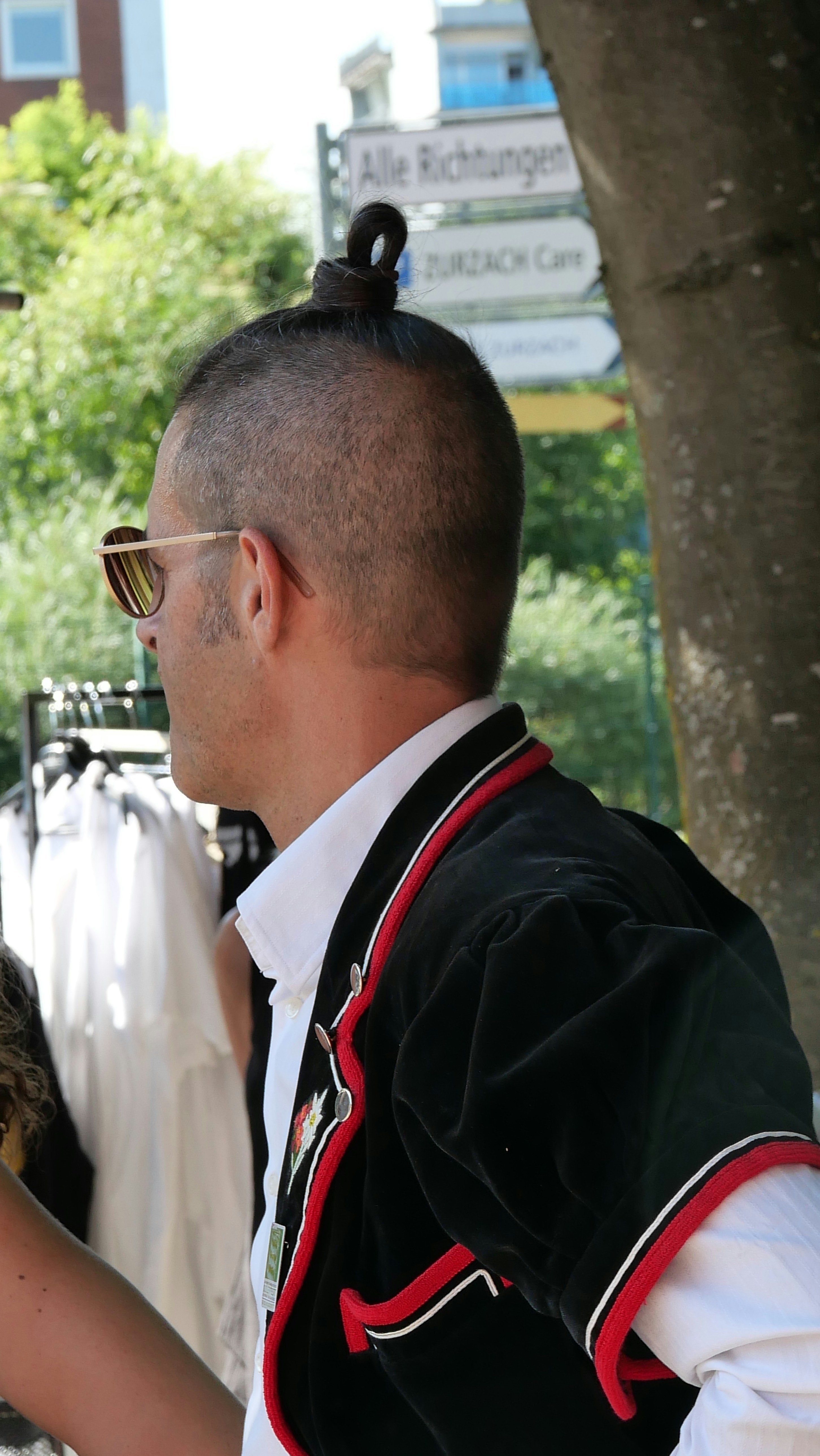 Portrait of a man in sunglasses, shown from the side, wearing a black jacket with red trim; background features a leafy street and a blurred sign. The focus is on the subject’s profile and attire.