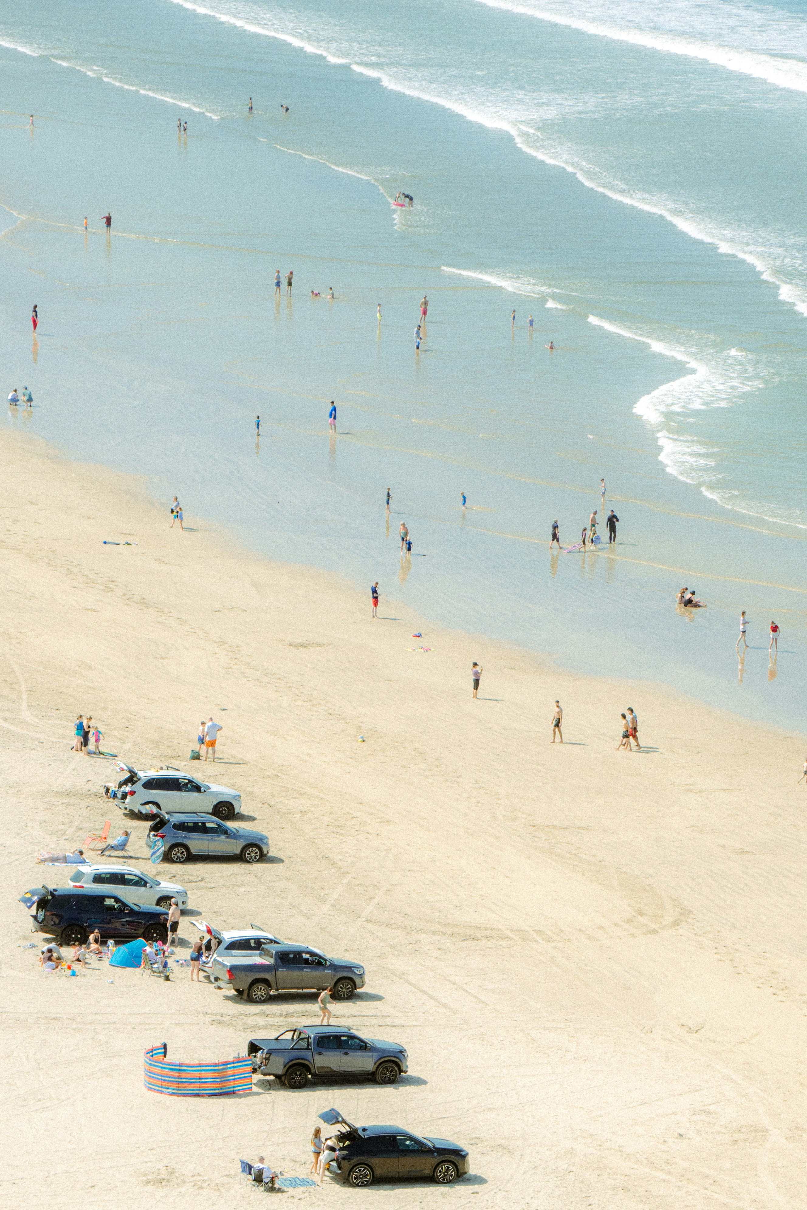 Sunlit Shoreline: Beachgoers, Cars, and Gentle Waves