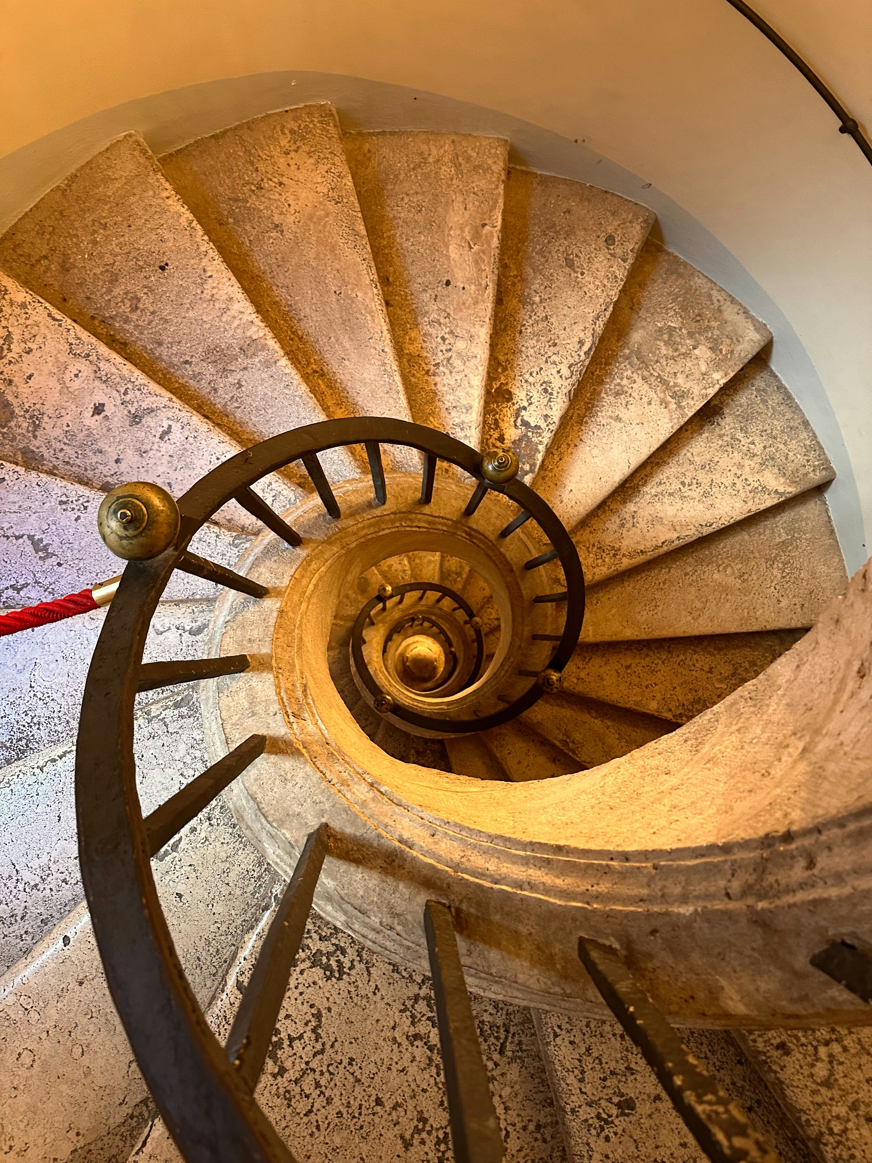 Coiled stone staircase descends in a tight spiral with warm amber lighting and iron railing accents.