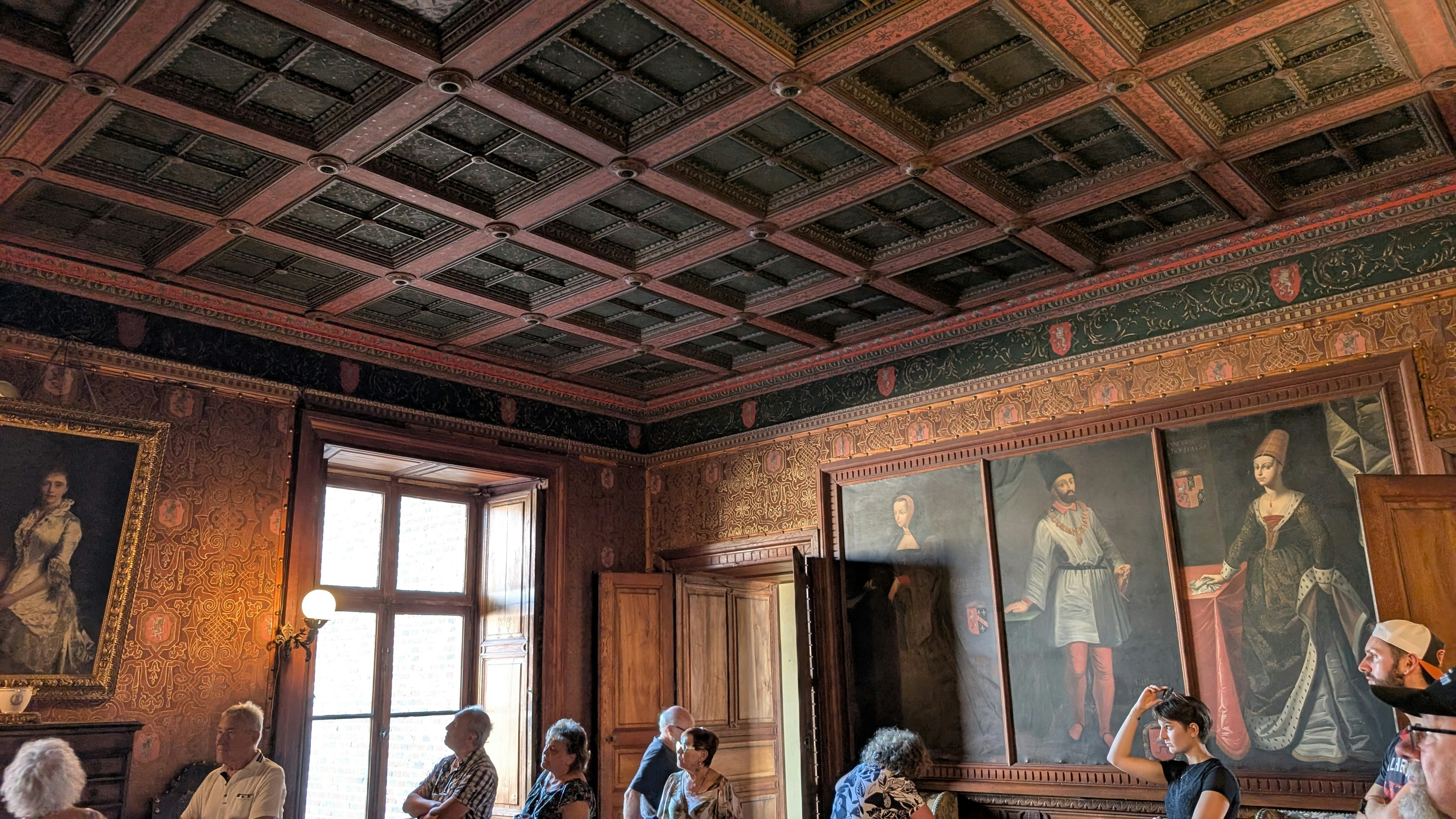 Historic interior with ornate coffered ceiling, gilded walls, and large portraits; visitors seated and observing art along a doorway-lit wall.