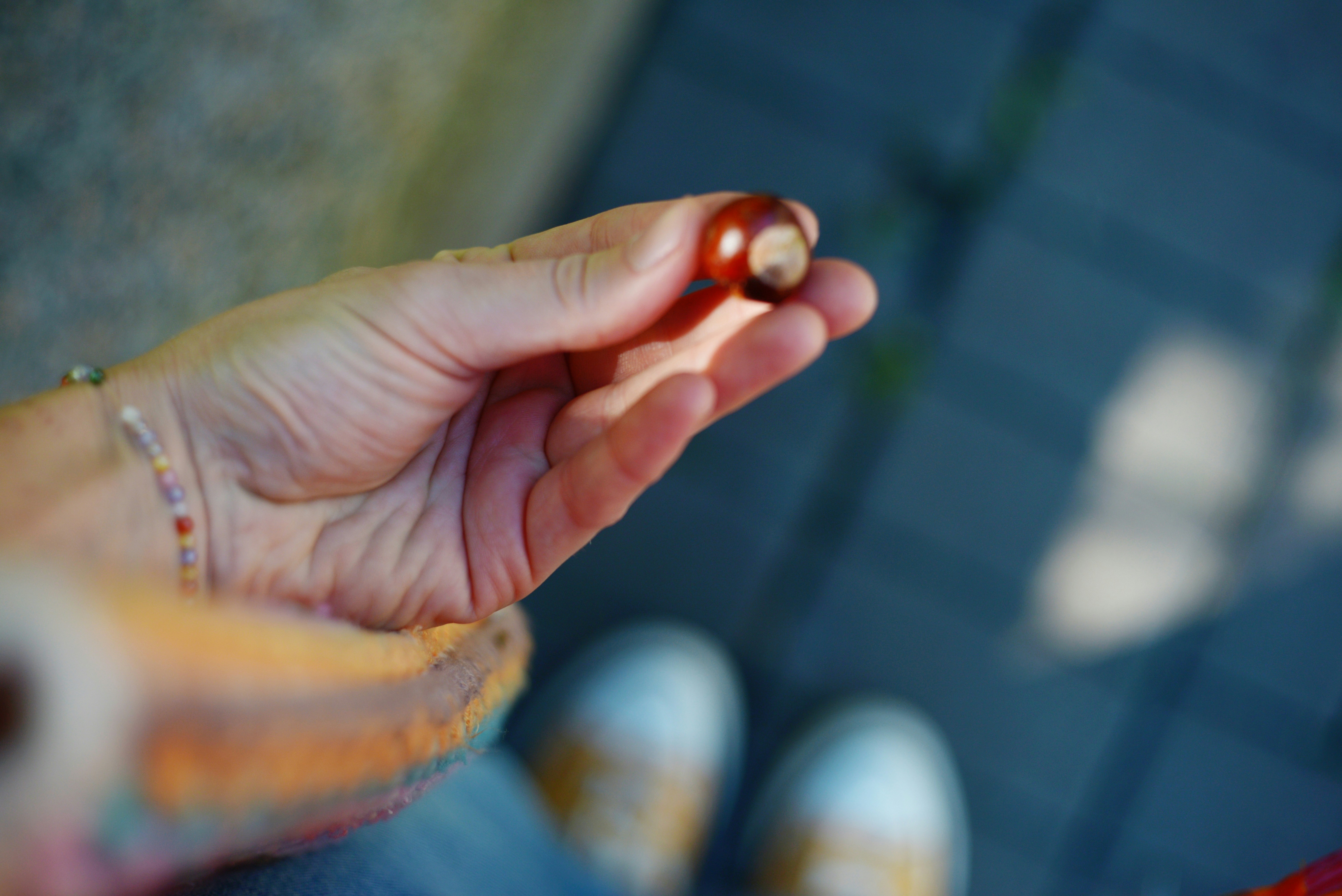 Close-up of a hand pinching a small cylindrical object with a blurred floor and shoes in the background. The scene highlights a personal, everyday moment with a focus on the object held.