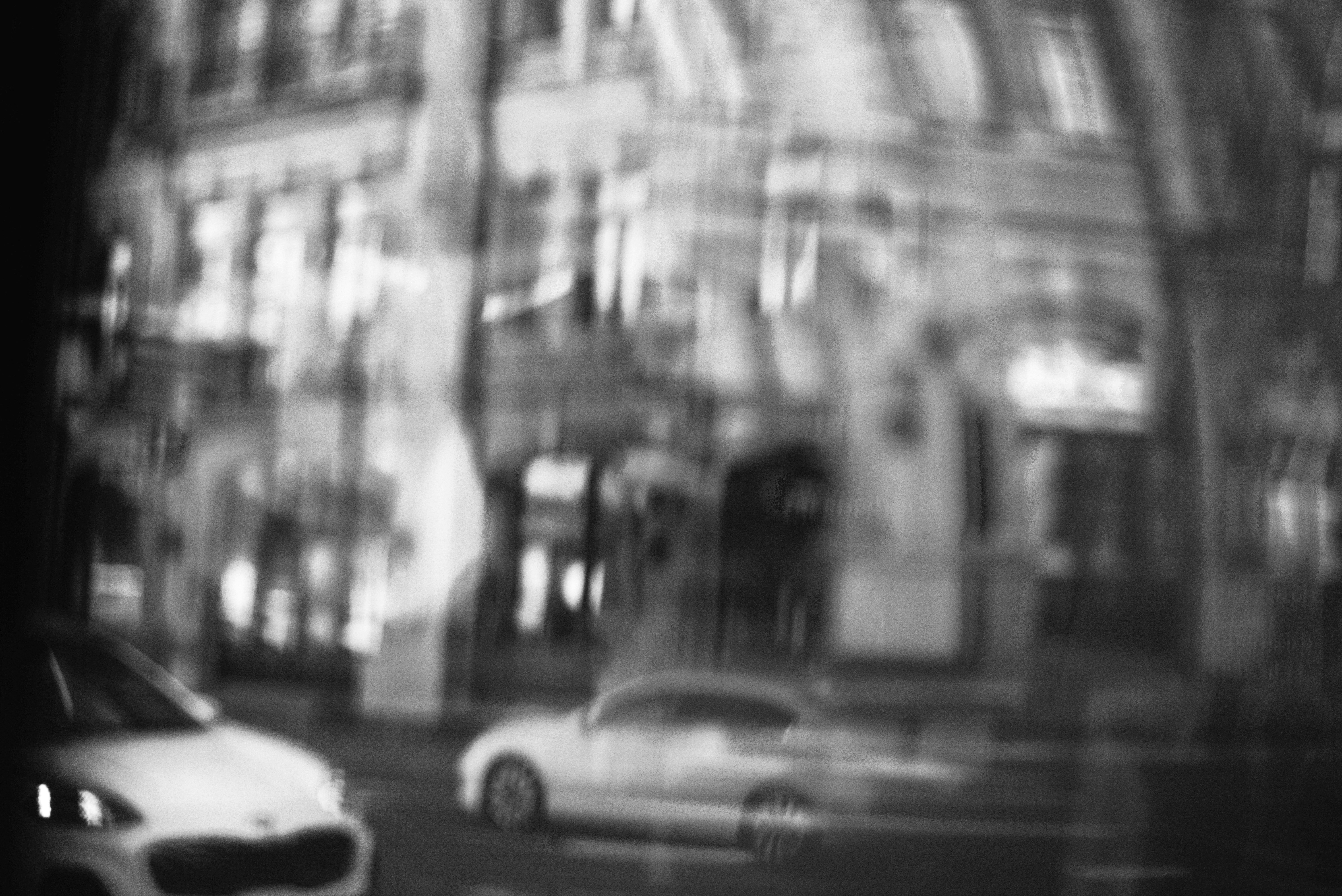 A blurred city street scene with cars and indistinct storefronts reflected through a glass surface, conveying motion and depth. This is a photographic capture with atmospheric distortion.