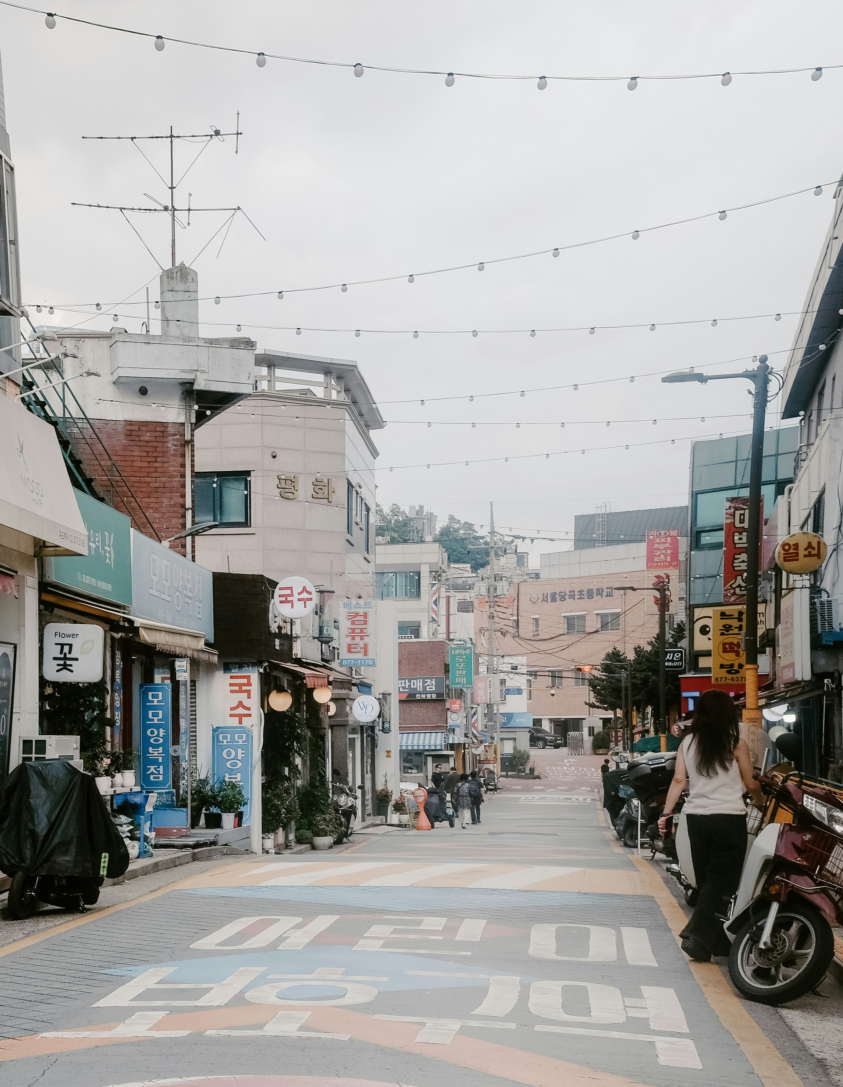Bustling urban street lined with small shops and signs, a pedestrian walks away from the camera; string lights overhead add a festive mood to a quiet daytime scene. (Note: not a painting or illustration.)