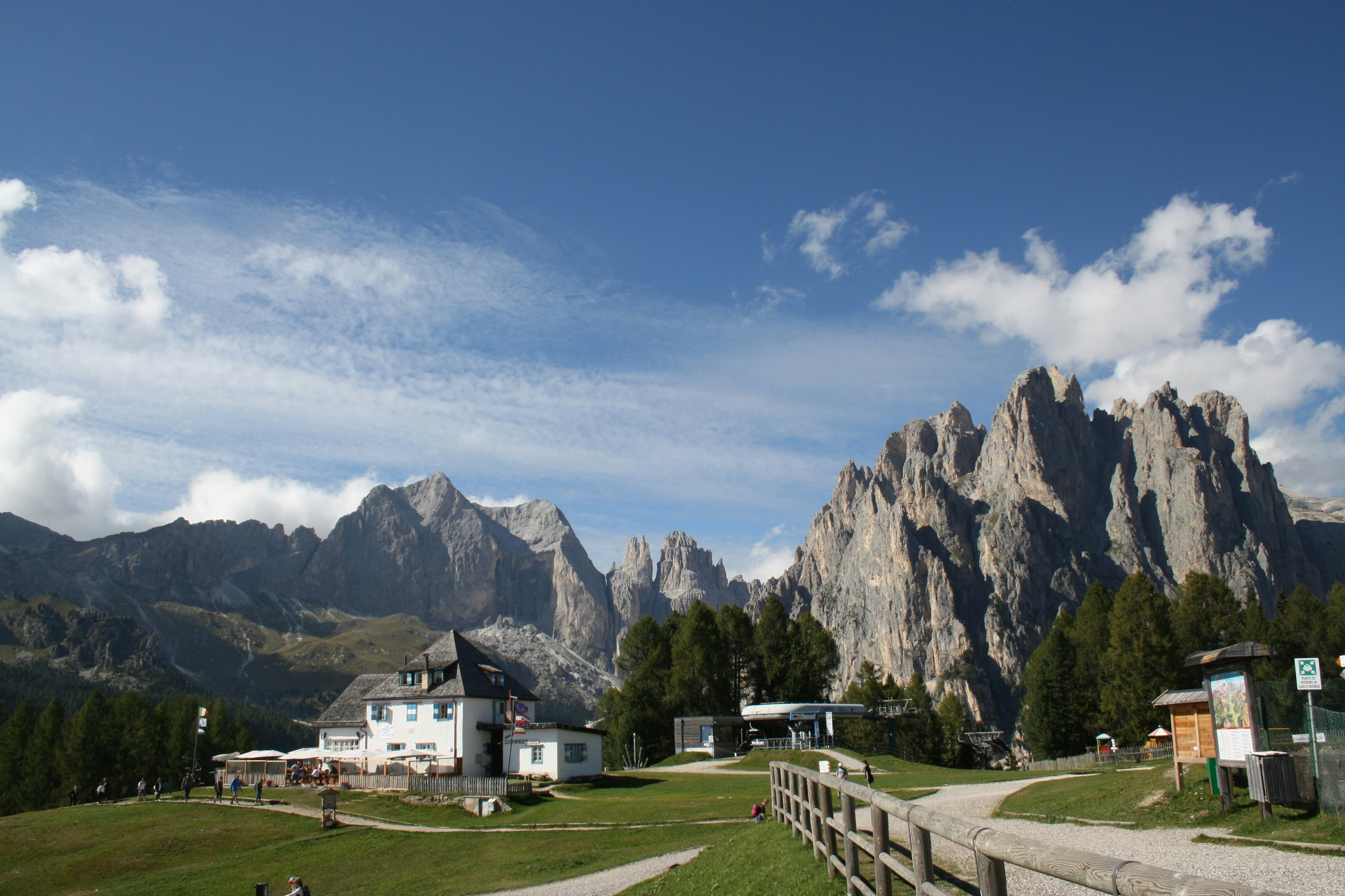 Alpine village buildings and a wooden fence sit near grassy slopes with towering jagged mountains in the background under a blue sky.