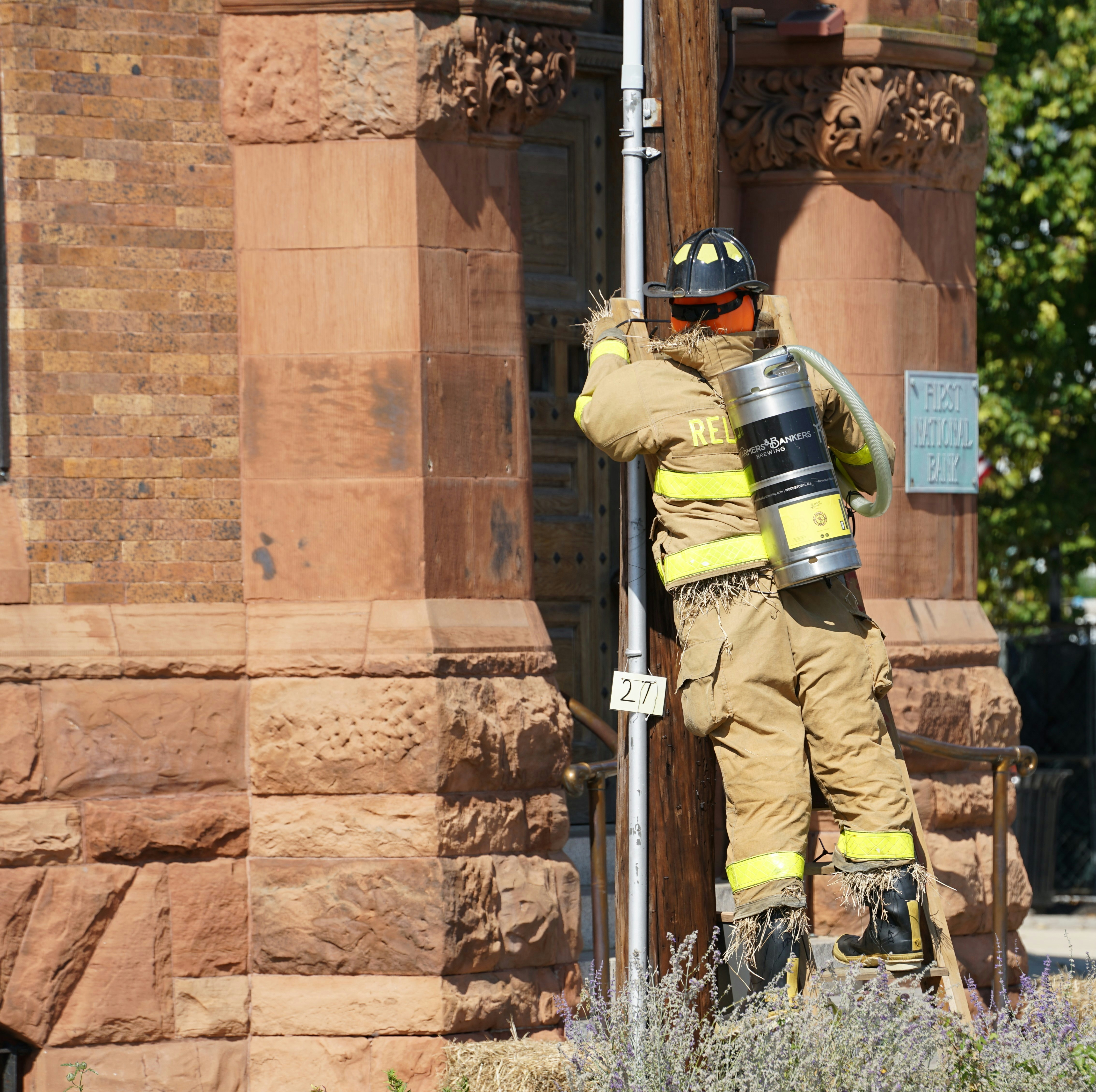 Firefighter in full turnout gear with an oxygen tank stands at a historic brick doorway, preparing to enter as a responder. The scene emphasizes gear and stance near a stone building.