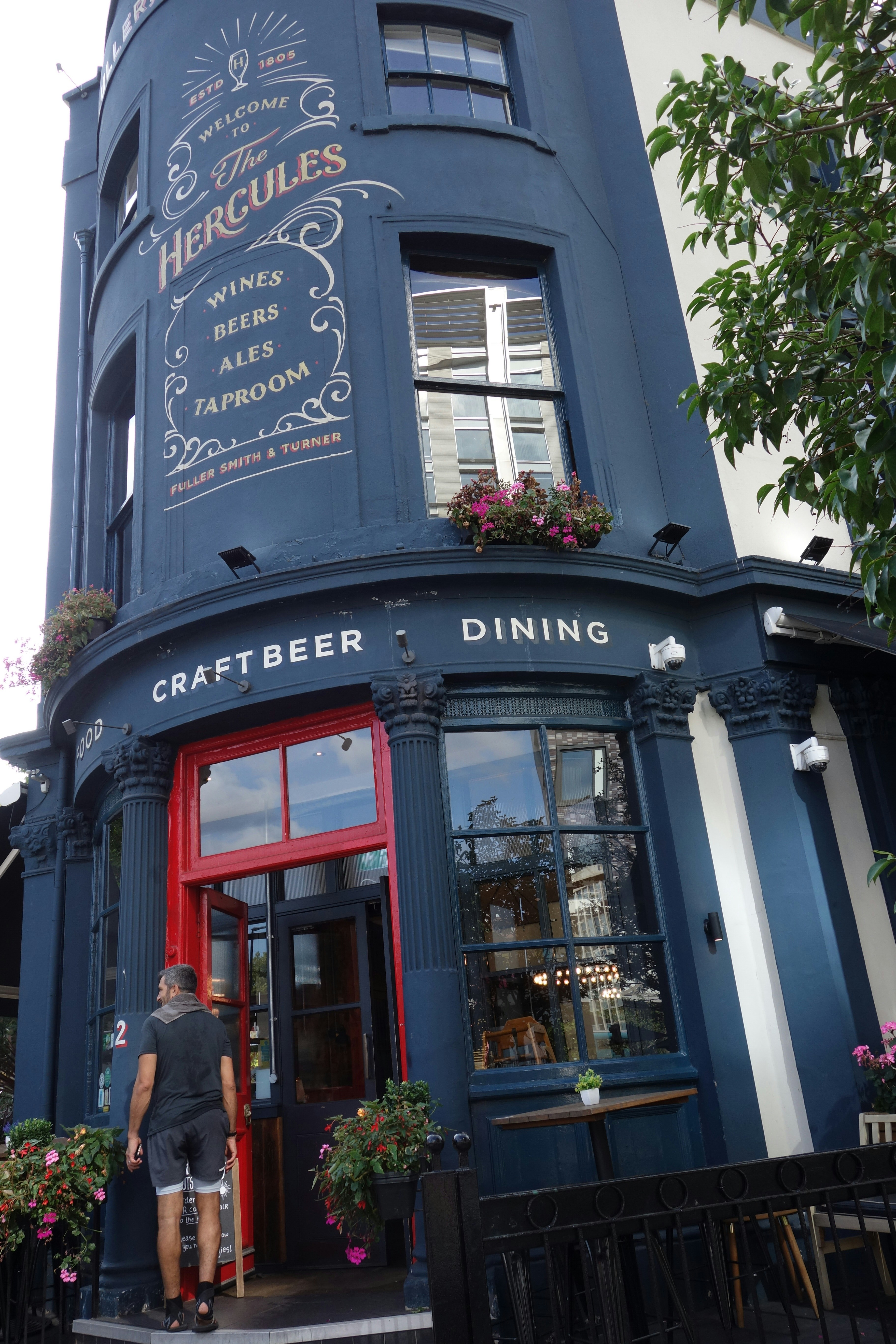 Blue-painted pub facade with ornate signage and a bright red doorway; a man stands at the entrance, plants in window boxes and outdoor seating visible.