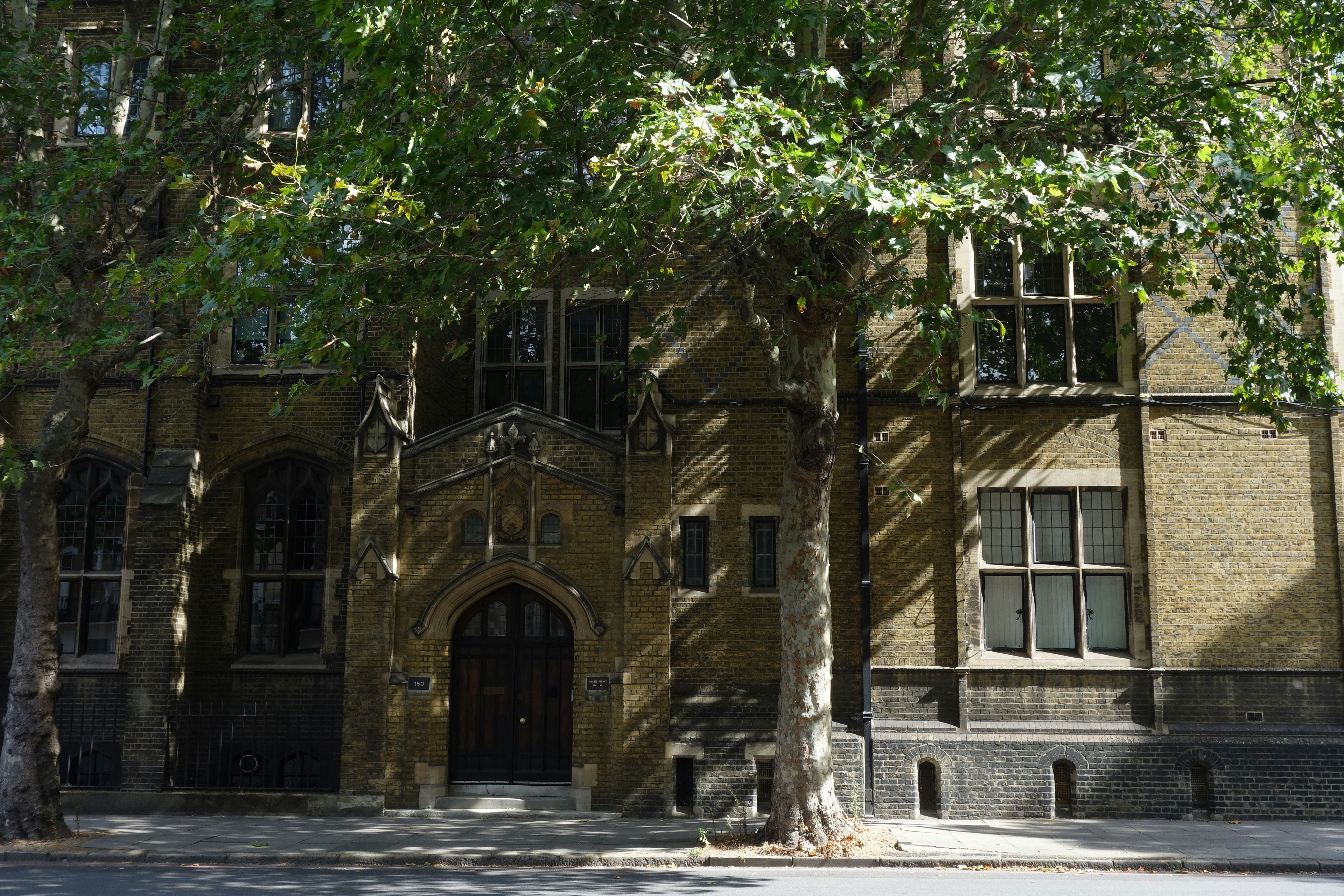 Brick building with arched doorway and large windows framed by mature trees along a sunlit sidewalk.