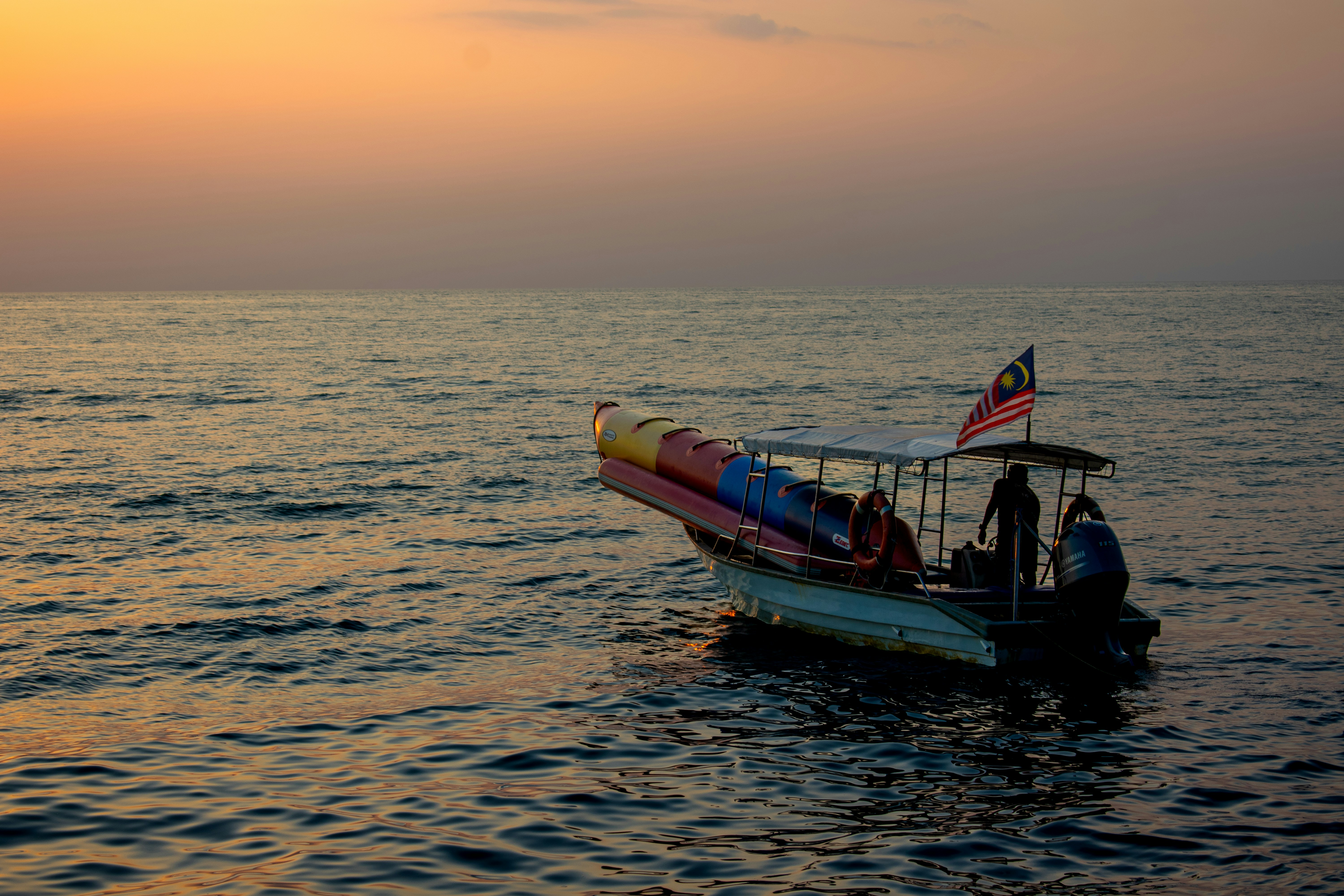 A small colorful boat drifts on tranquil sea at sunset, Malaysian flag fluttering as the warm sky bleeds into the water. This is a photography scene of a seafaring craft against a serene horizon.