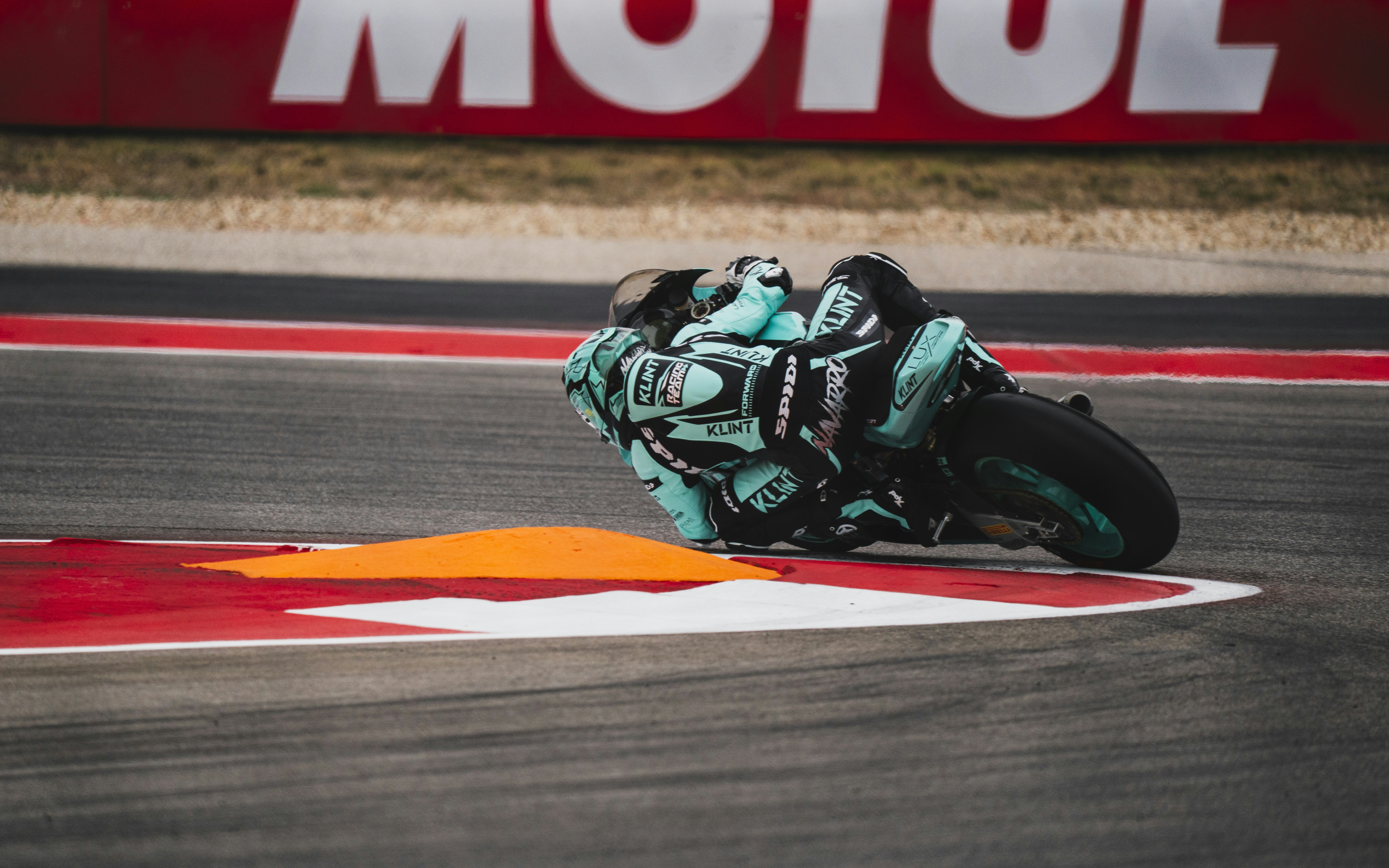 A teal-marine race motorcycle leans through a sharp corner on a track, rider tucked and aerodynamic, with bold track markings and a blurred red signage backdrop.