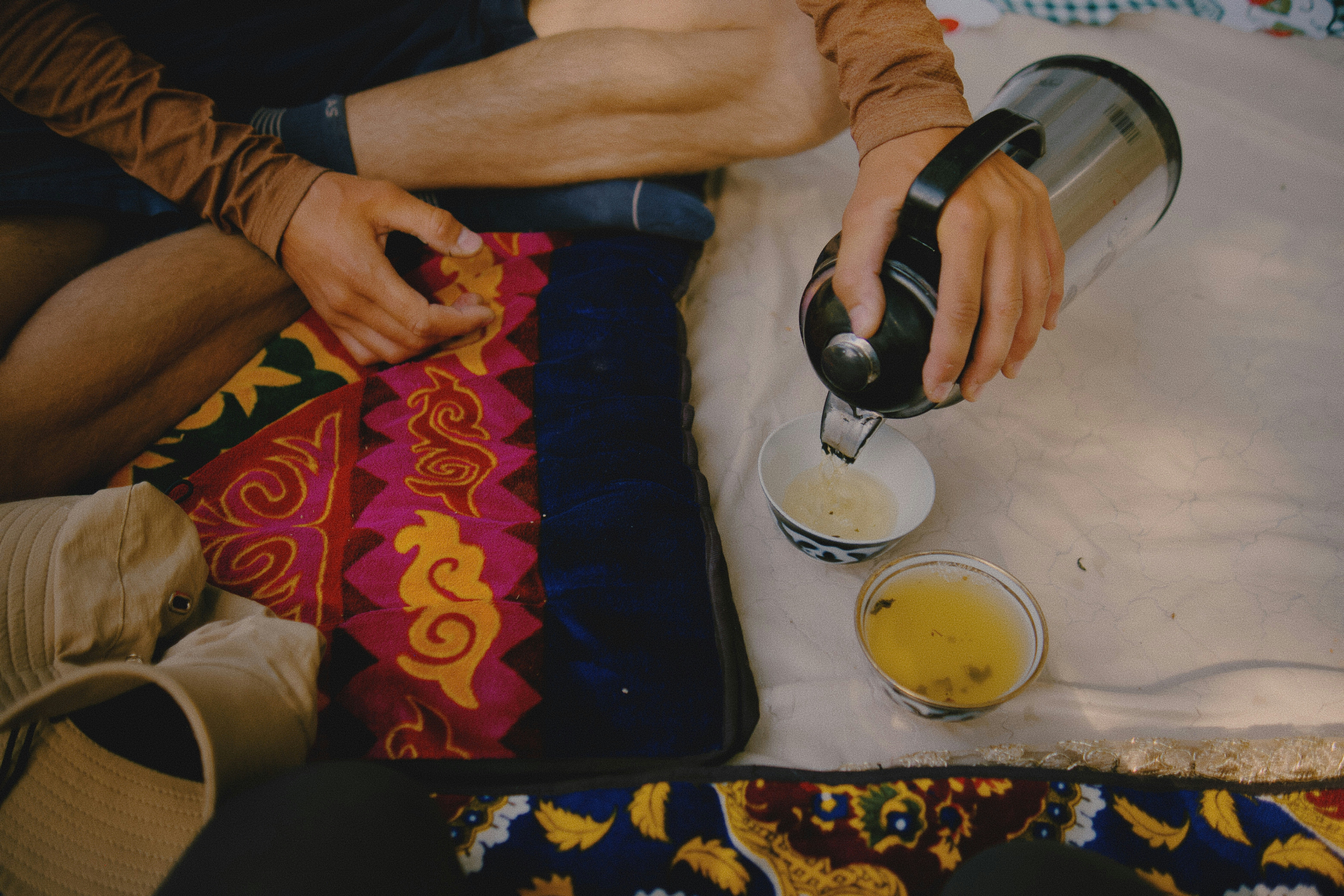 Close-up of a person pouring a beverage from a thermos into a small cup, with vibrant textile and a second drink nearby on a metal surface.