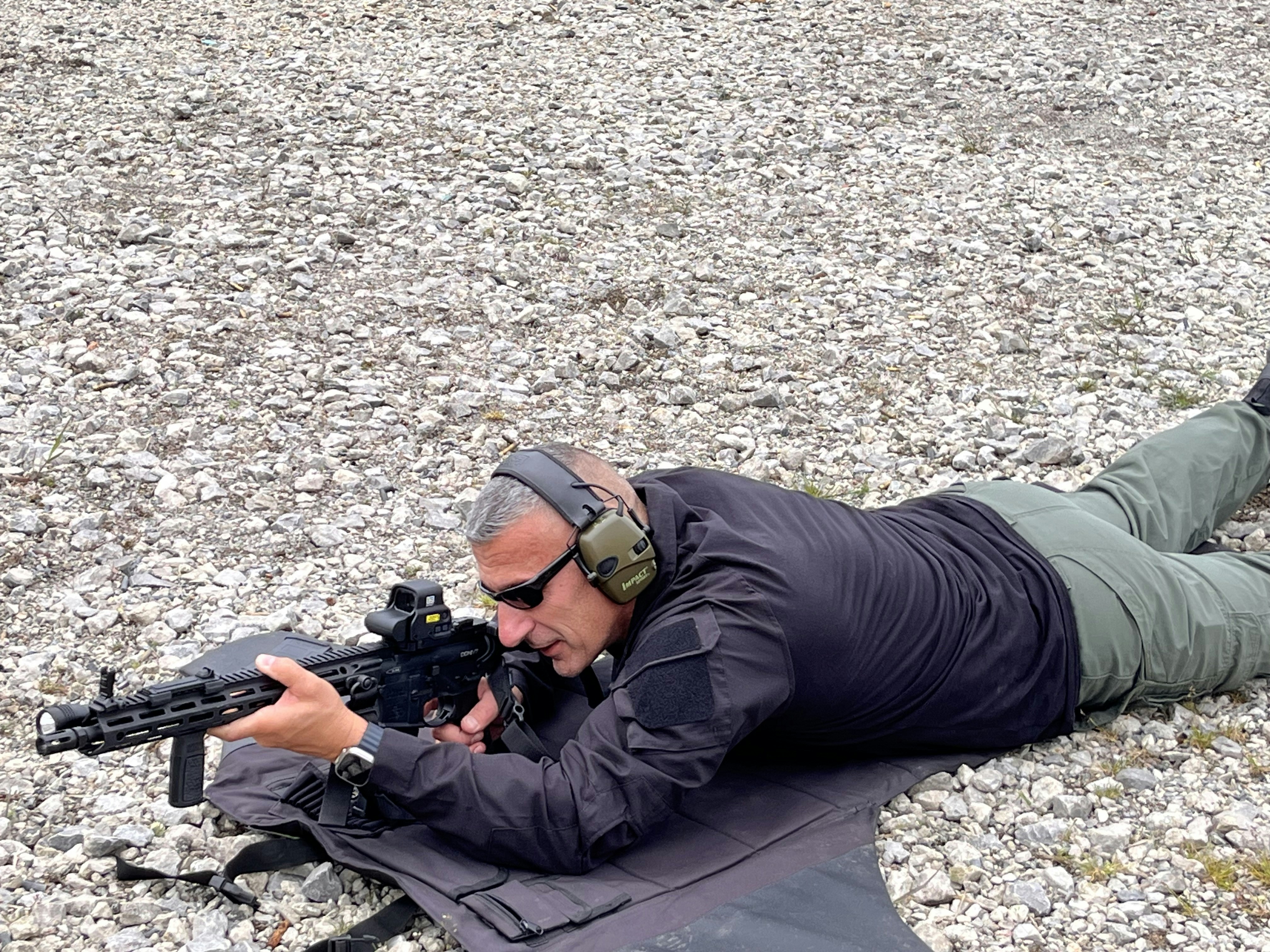 A prone shooter stabilizes on a gravel surface with a modern rifle and earmuff protection, aiming down sights in an outdoor range. A compact scene showing focus on the firearm and stance.