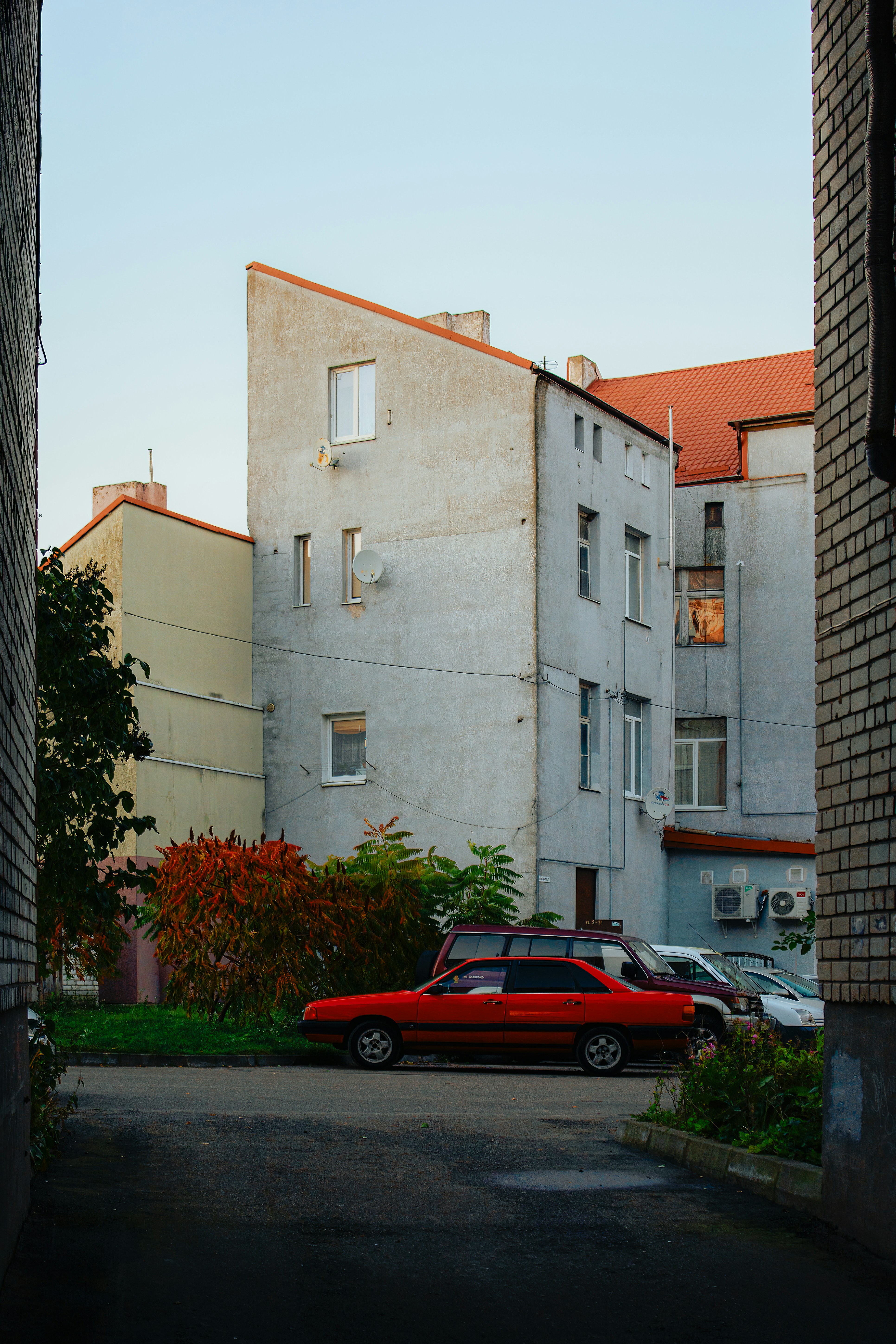 Red vintage car parked in a residential courtyard with pastel apartment buildings in the background, quiet afternoon light on a calm street.
