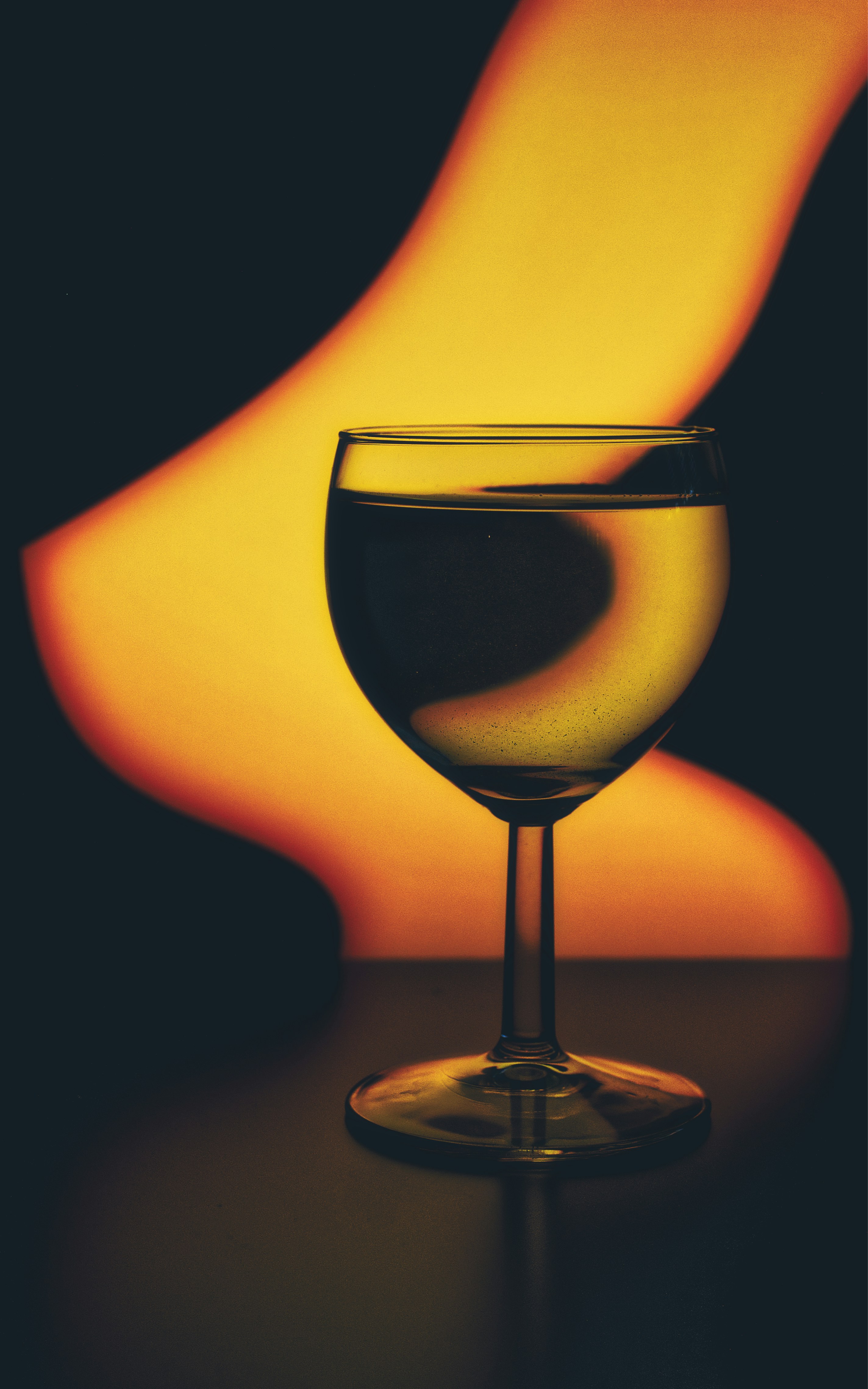A glass of water stands on a dark surface, framed by a bold, curved orange glow that adds dramatic backlight and abstract warmth to the composition.