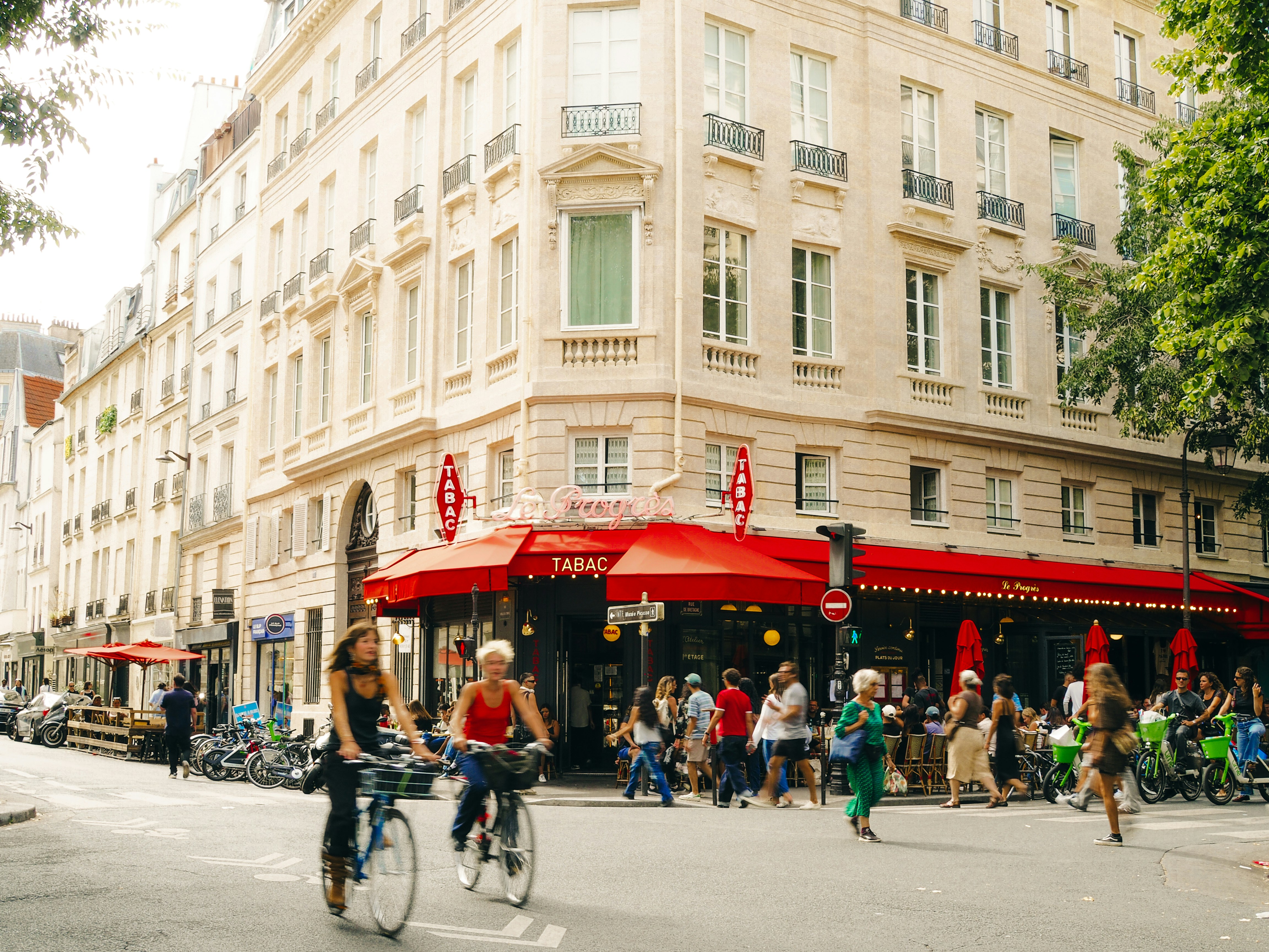 Bustling street scene with a red-awning café at a historic Parisian corner, pedestrians and cyclists in motion under warm daylight.