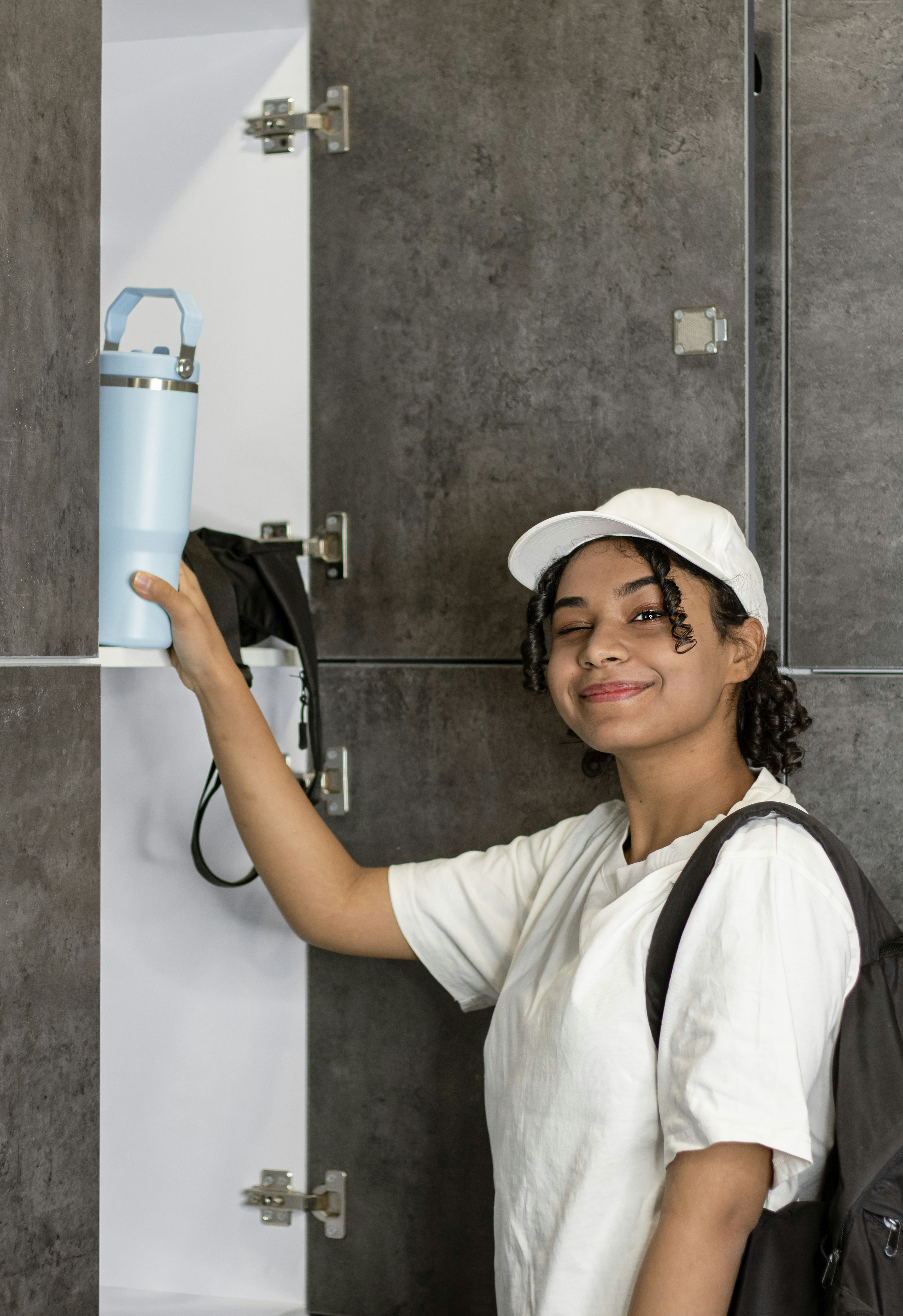 A cheerful student stands beside an open locker holding a light blue water bottle, wearing a white cap and t-shirt with a backpack slung over one shoulder.