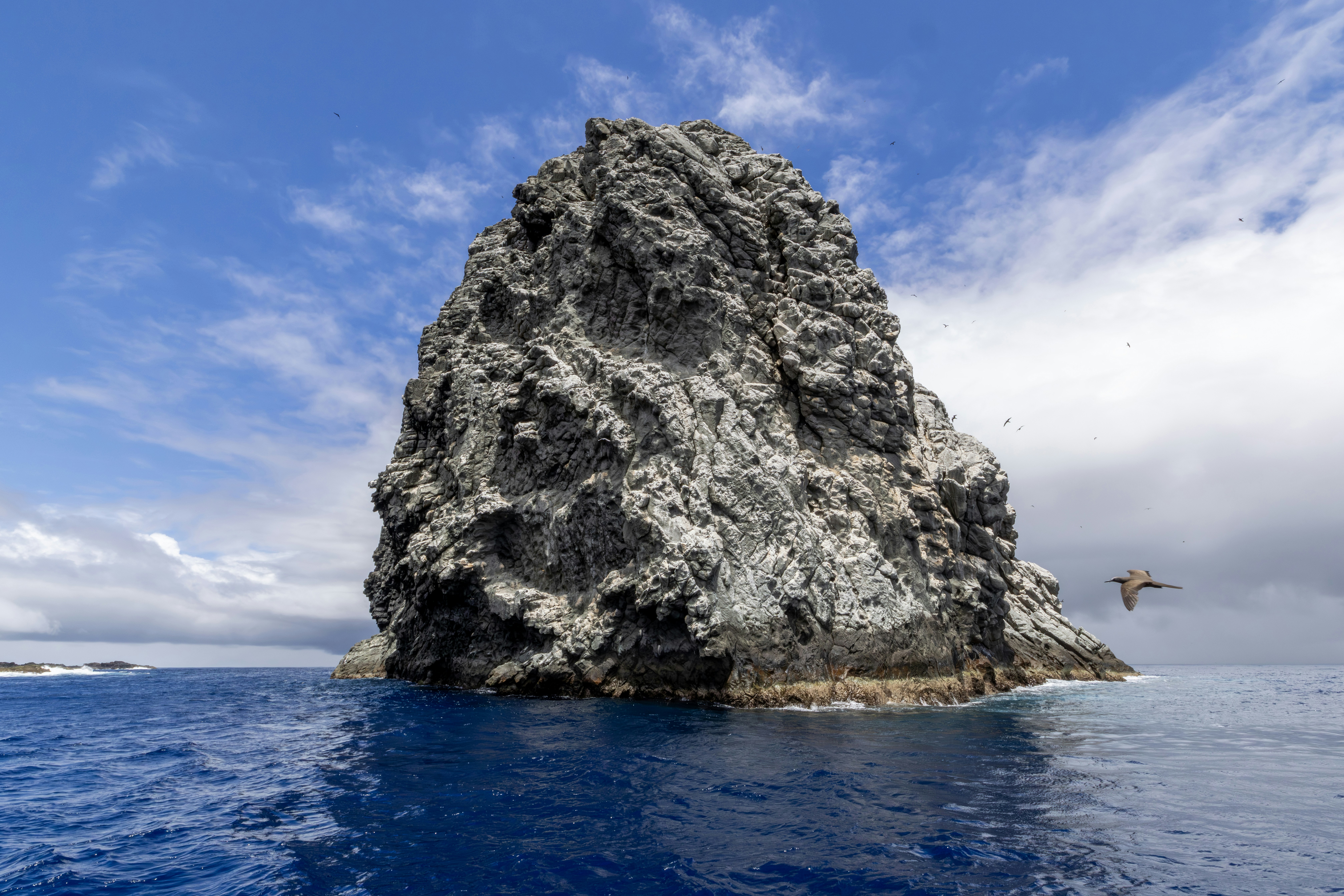 A solitary rugged rock formation emerges from the sea against a blue sky, with a distant bird in flight near the waterline.