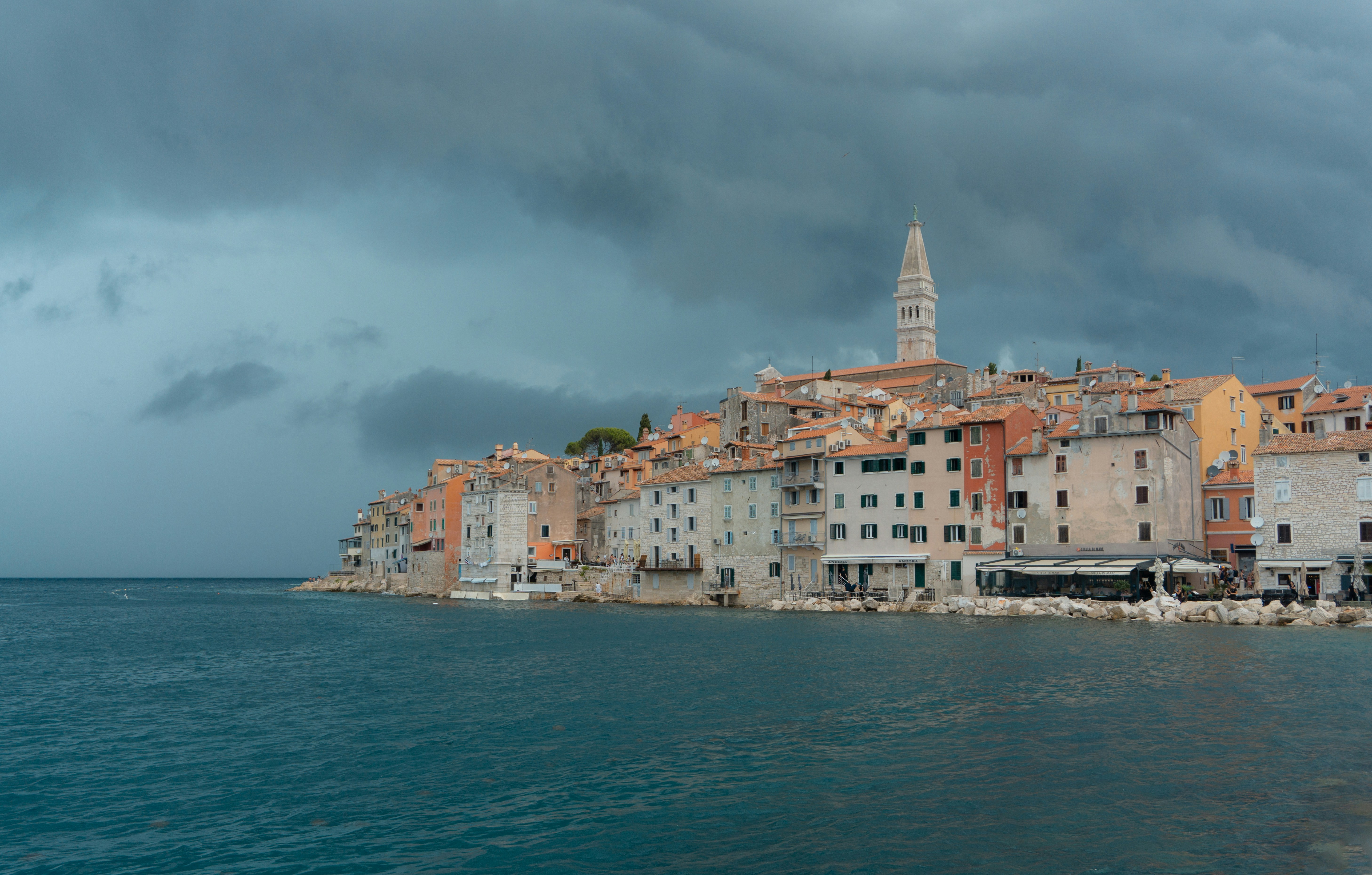 Colorful coastal houses cluster along the shore with a prominent church bell tower rising above, meeting the calm sea under a moody sky.