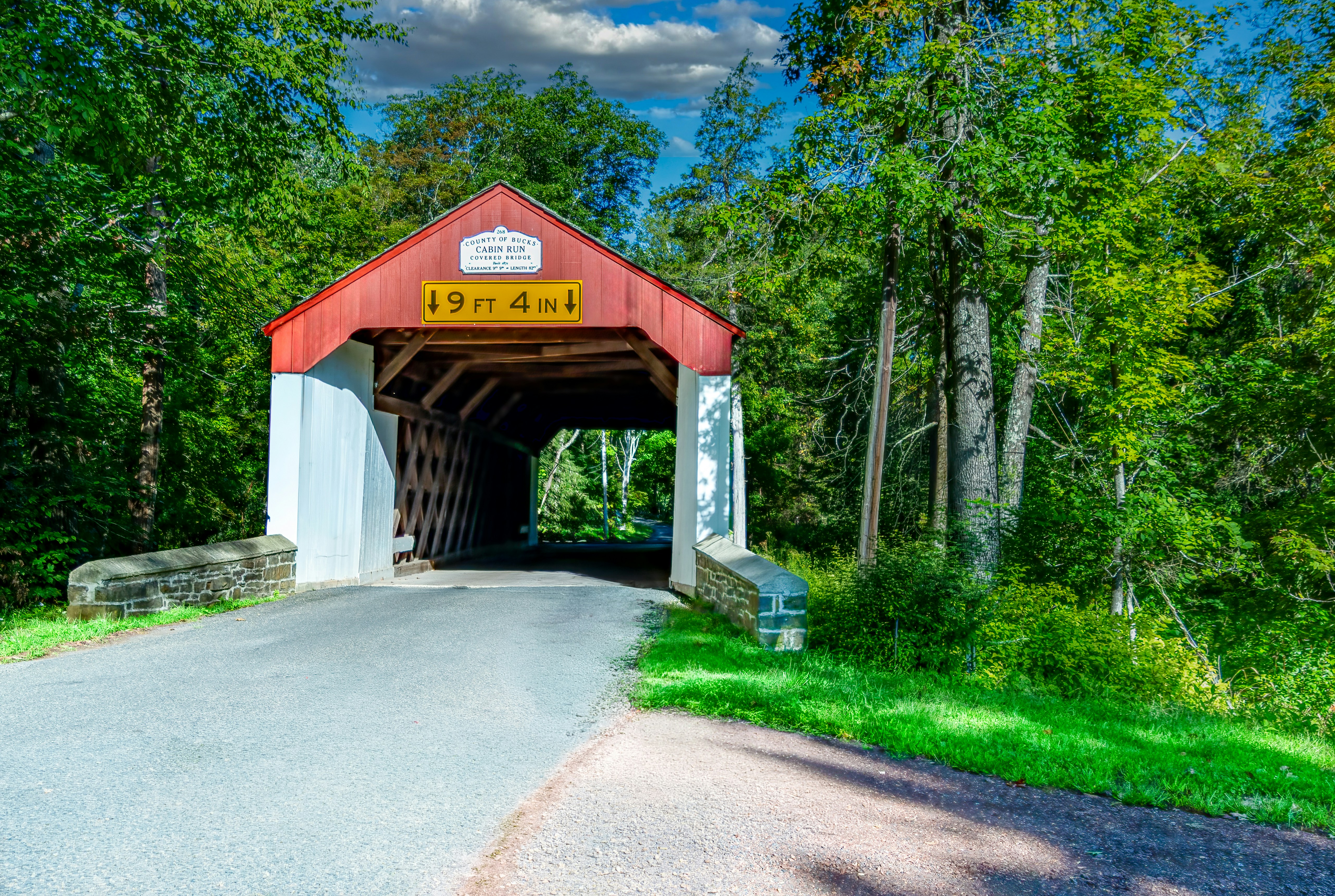 Red covered bridge framed by green trees and a paved approach, inviting a journey through the shaded timber tunnel.