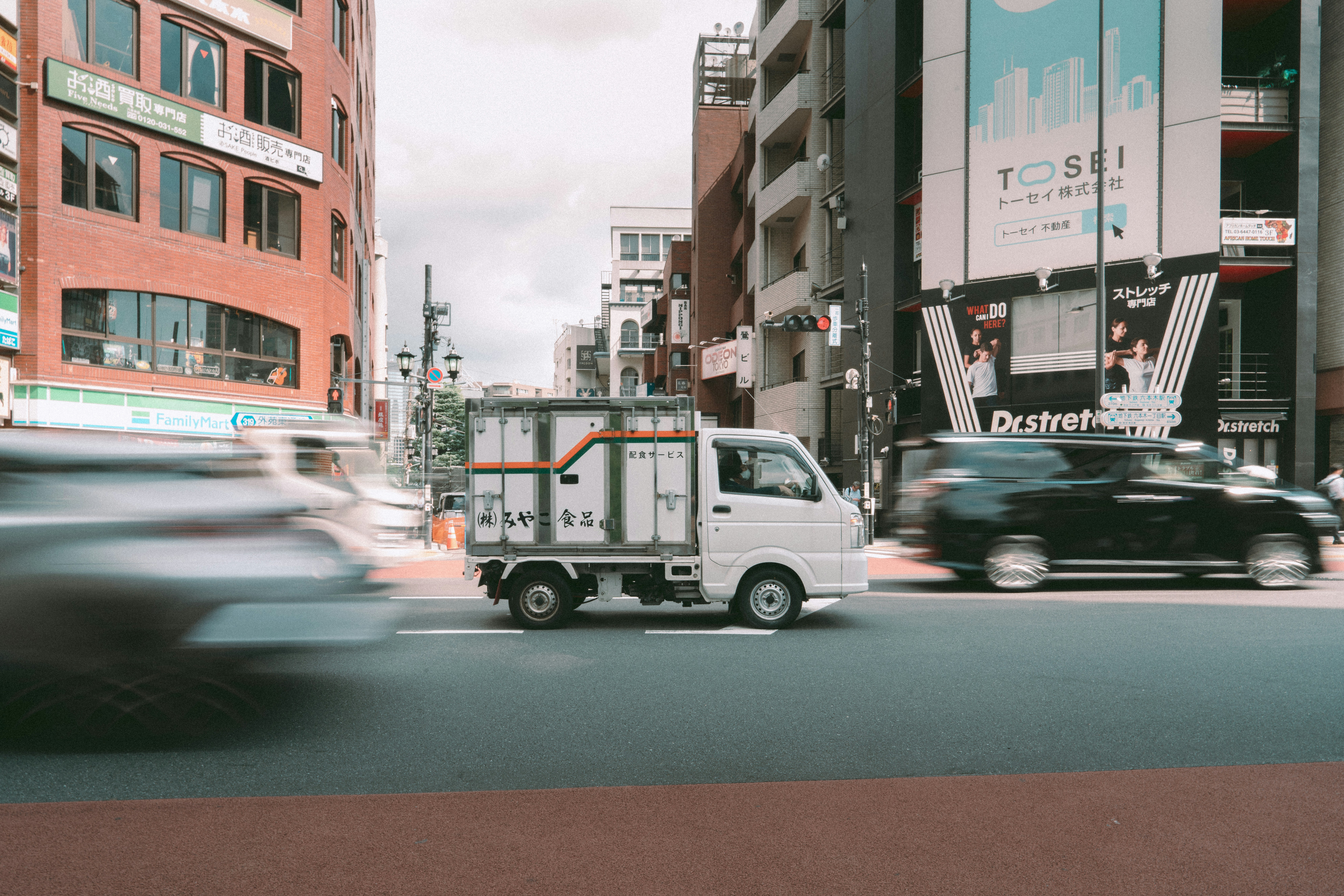 A white delivery truck moves through a bustling city street, blurred surrounding vehicles conveying speed and urban energy. The scene captures motion and layered storefronts in a shallow depth of field.