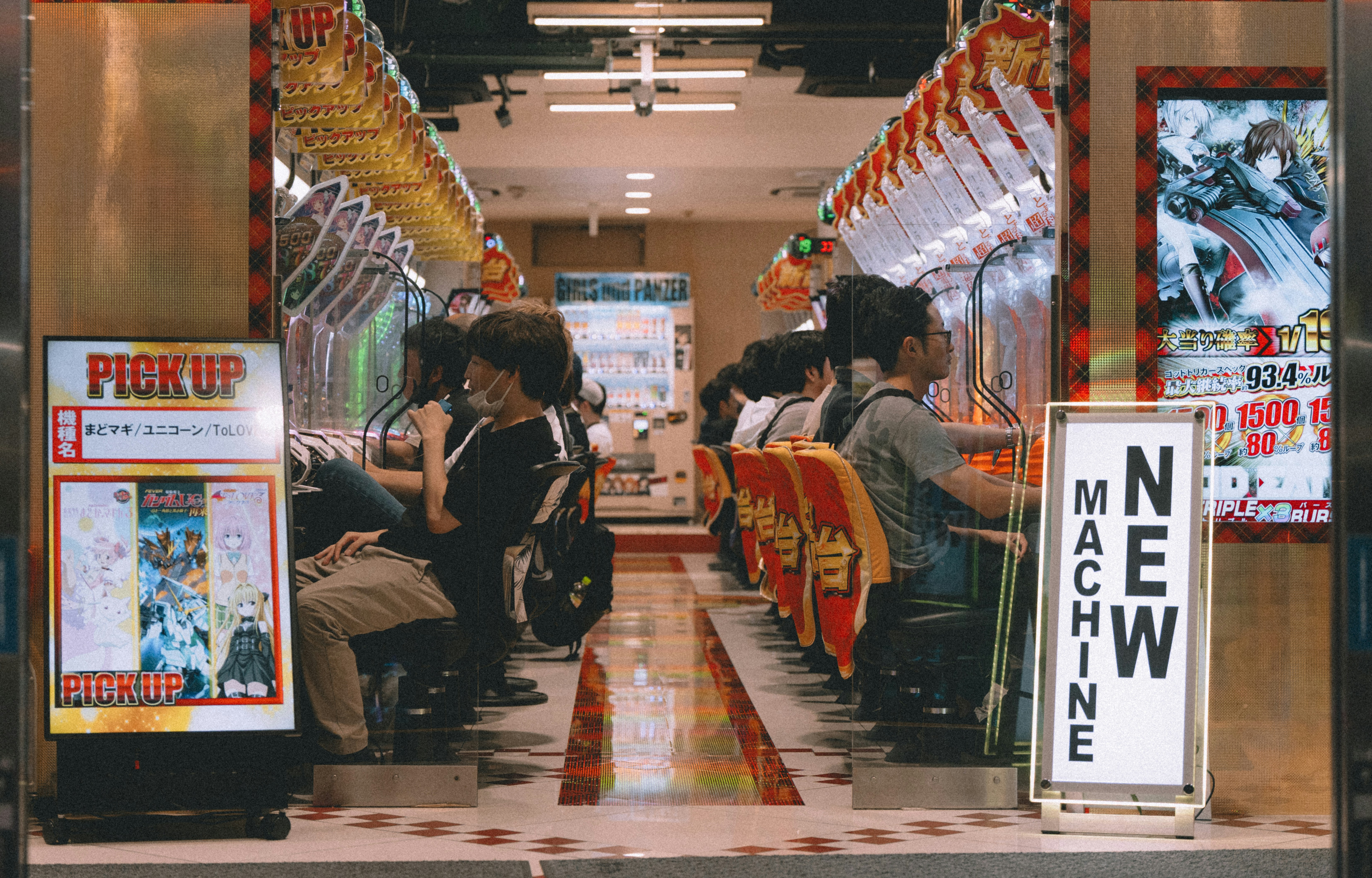 A busy arcade corridor with rows of claw machines on either side and players seated, awaiting their turn; signage and illuminated displays emphasize a lively gaming environment.