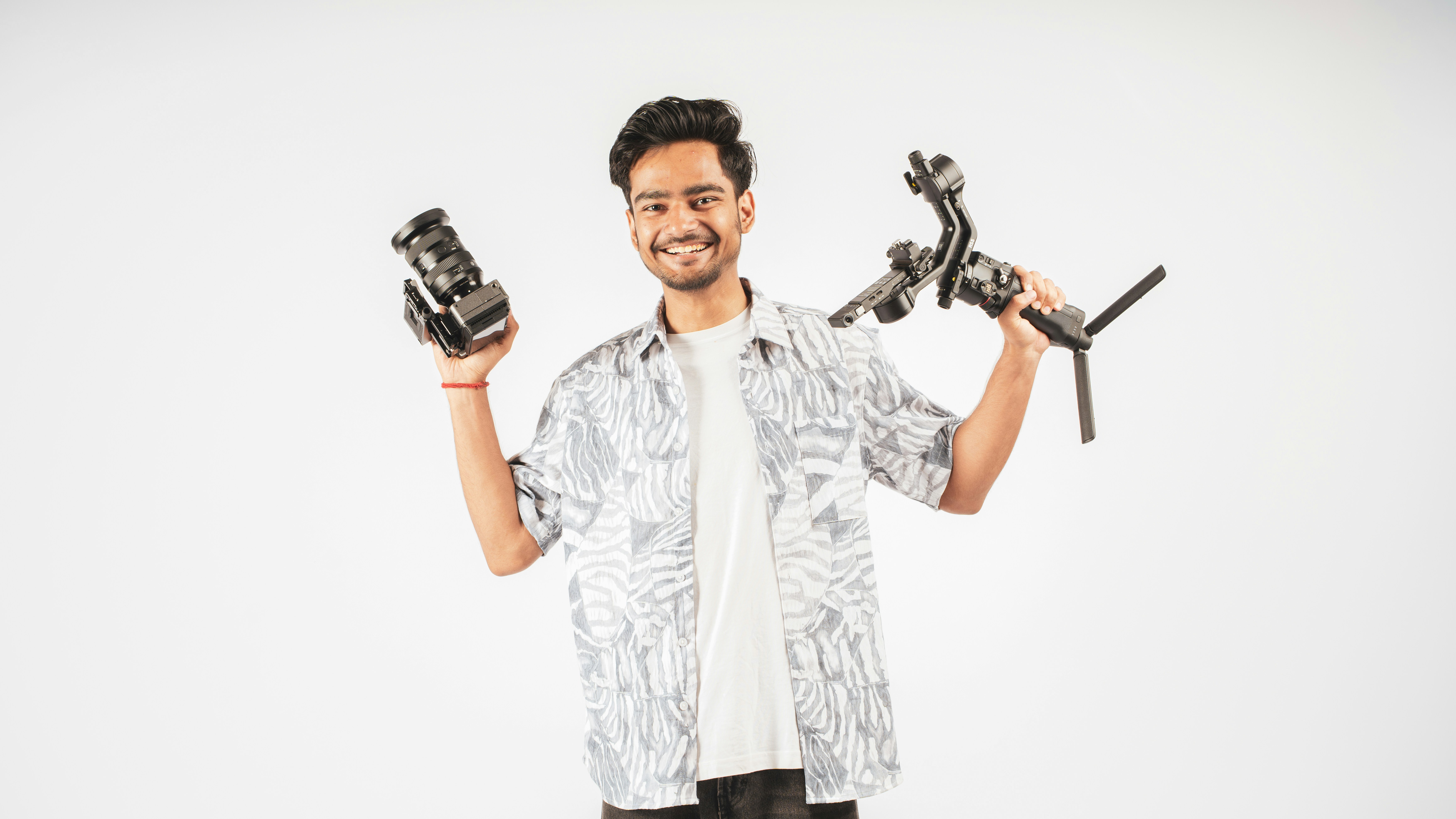 Cheerful man holds a camera and a drone gimbal on a bright, neutral backdrop, suggesting a talent for videography gear. This is a lifestyle/tech portrait.