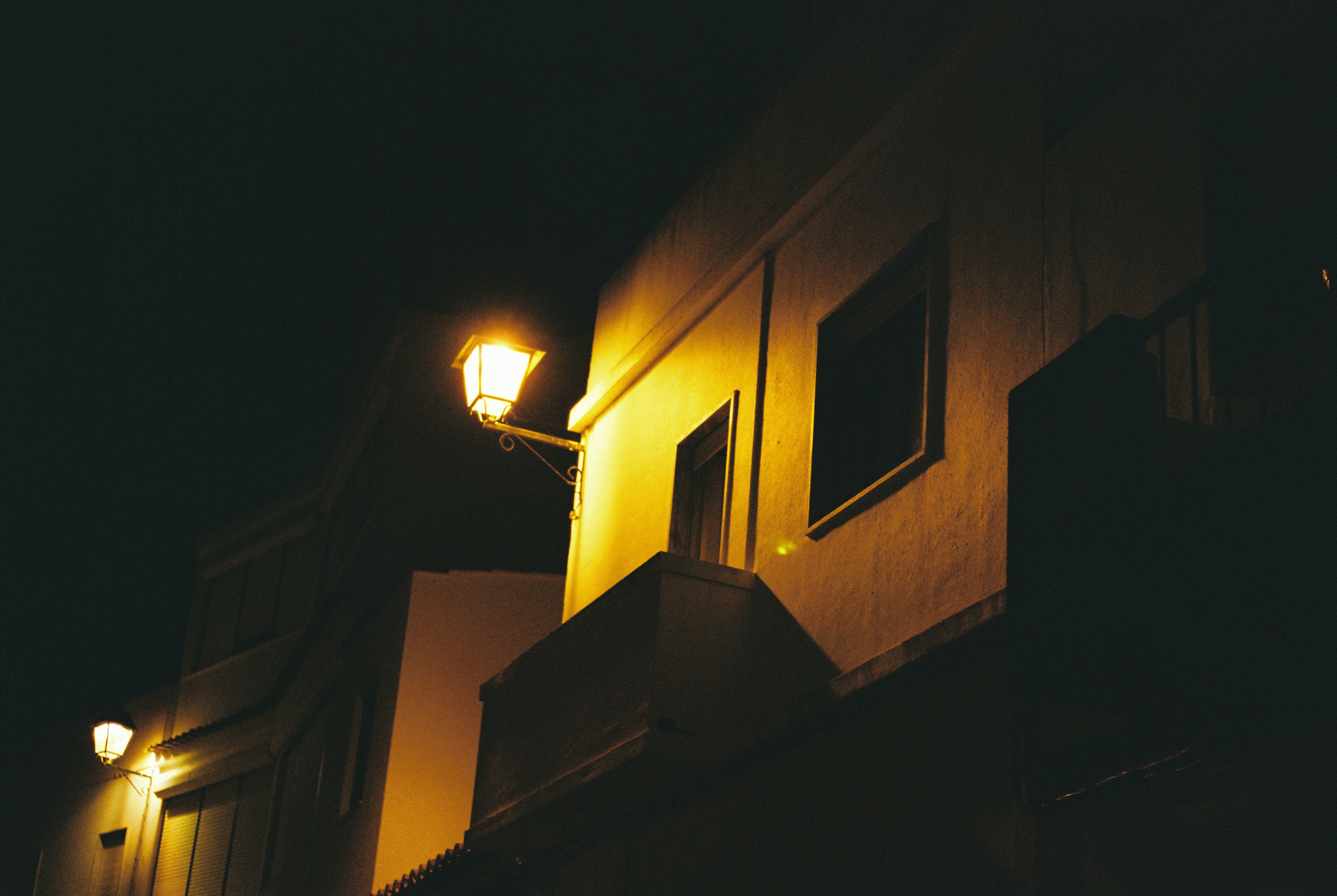 Warm streetlamps illuminate a quiet building facade at night, highlighting architectural edges and soft shadows. A moody, urban scene with subtle color and grain.
