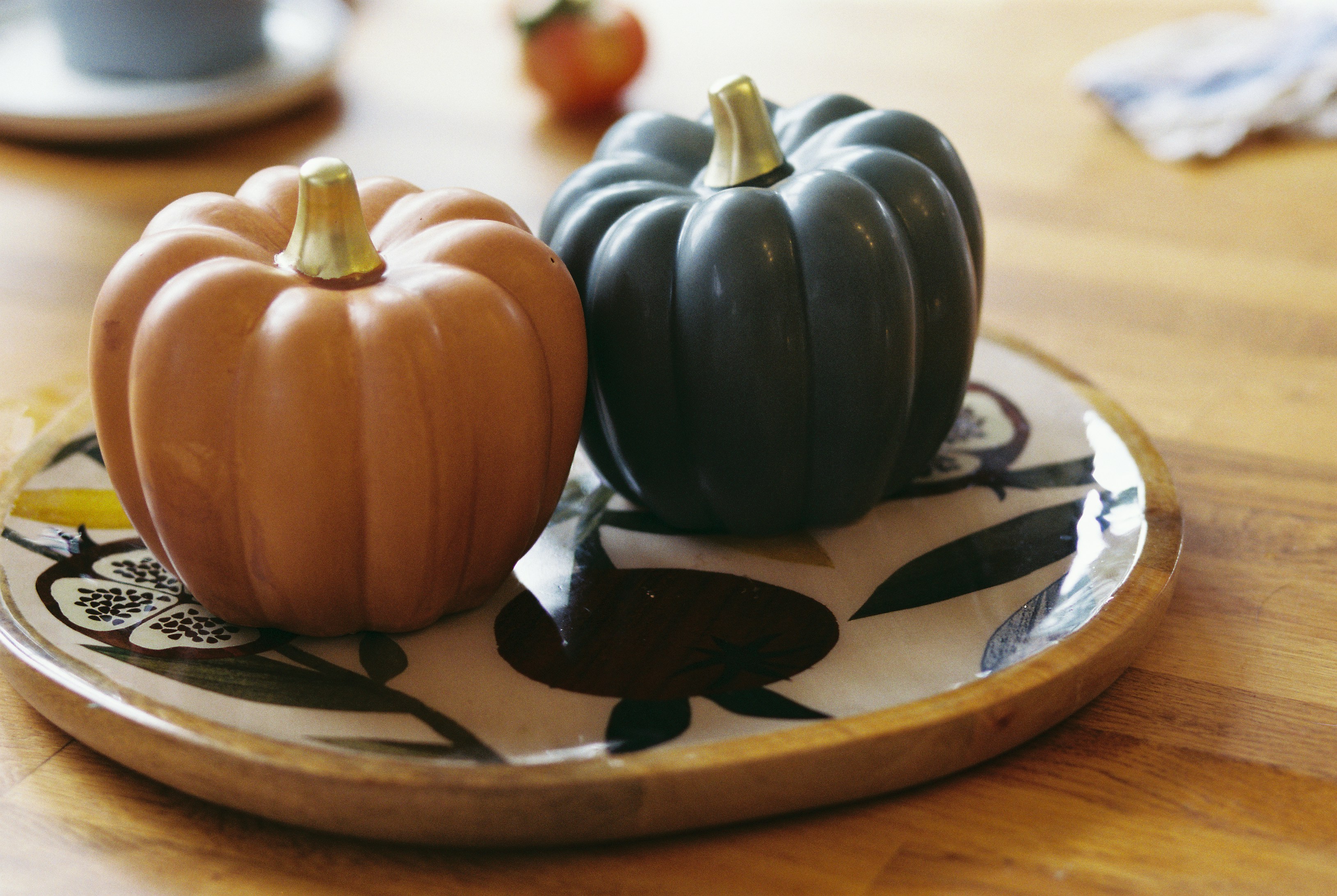 Two pumpkins sit on a patterned plate atop a wooden surface, with warm indoor lighting emphasizing their textures.