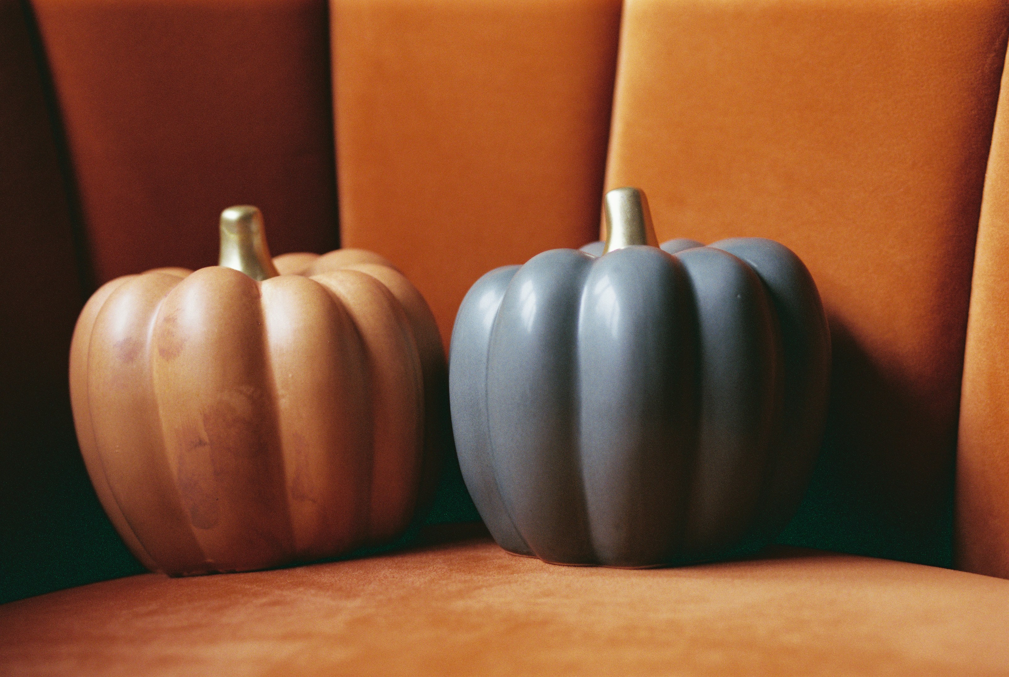 Two carved pumpkins resting on a plush orange seat with a soft, moody lighting and warm tones. Painted surfaces give a subtle, painterly feel to the scene.