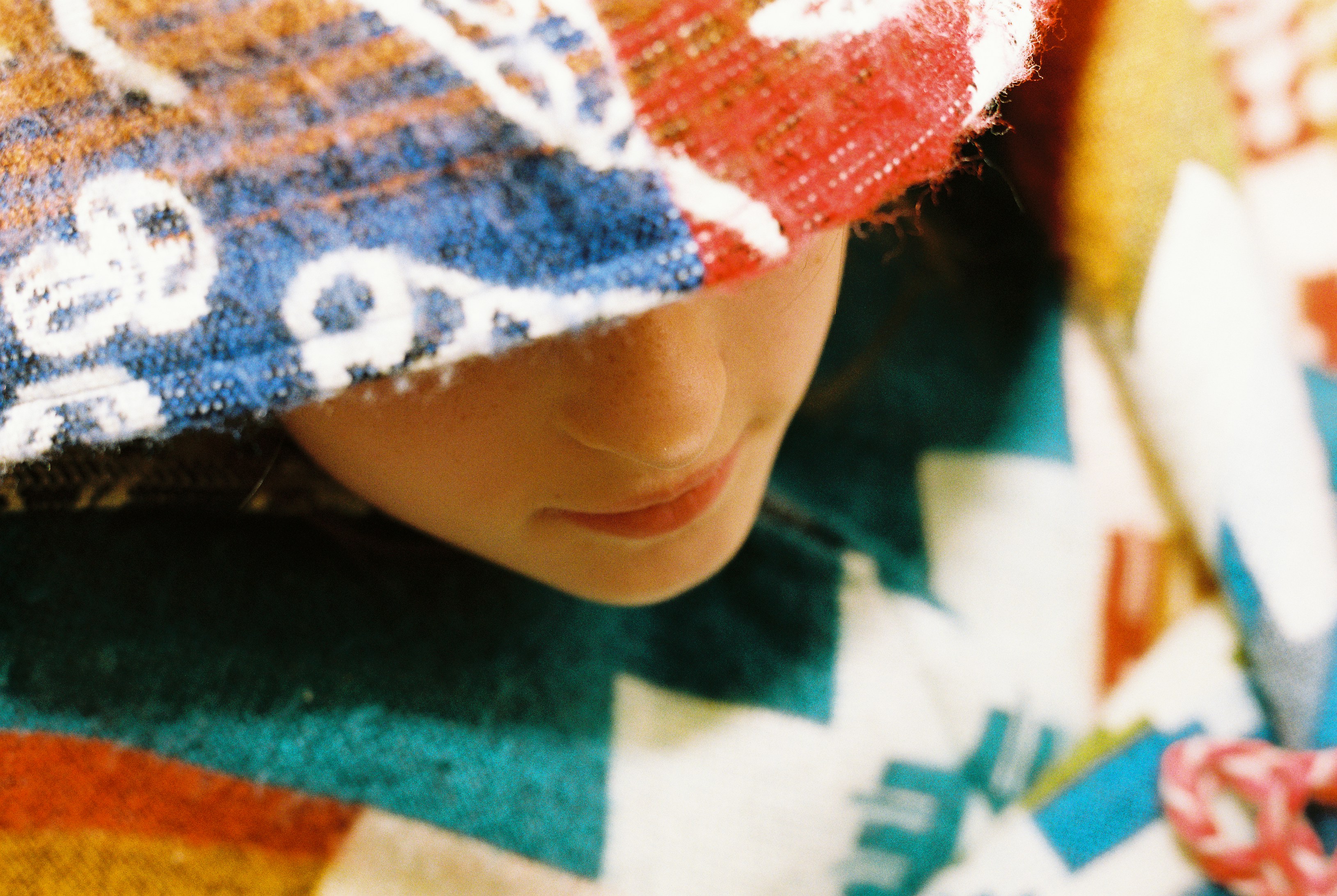 Close-up of a child’s lower face peeking from a vibrant patterned cloth, emphasizing the soft lips and colorful textile surrounding it. A photographic detail shot focusing on mood and texture; not a logo or illustration.