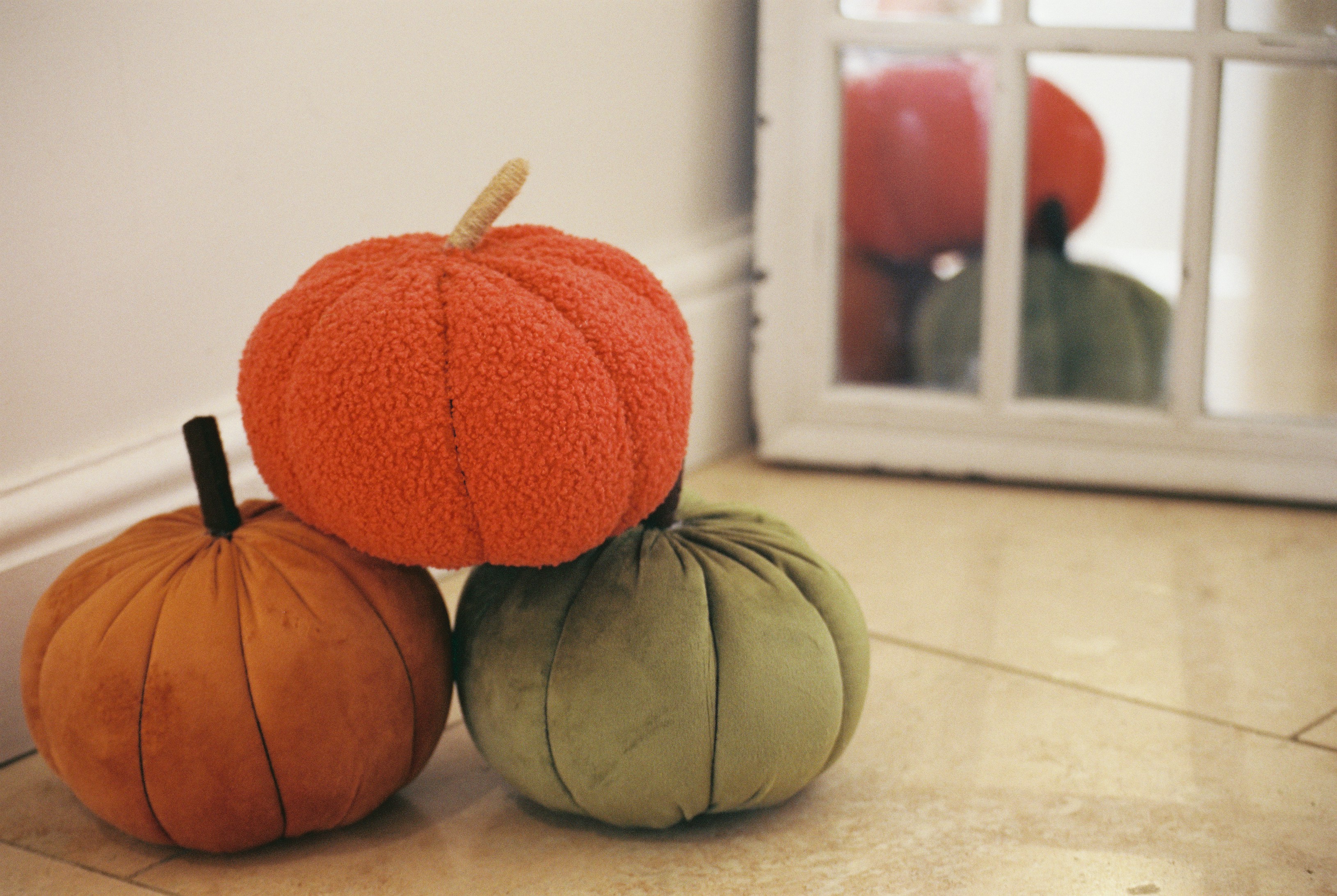 Three plush velvet pumpkins in orange, burnt sienna and sage green are stacked on a tiled floor beside a paned mirror.