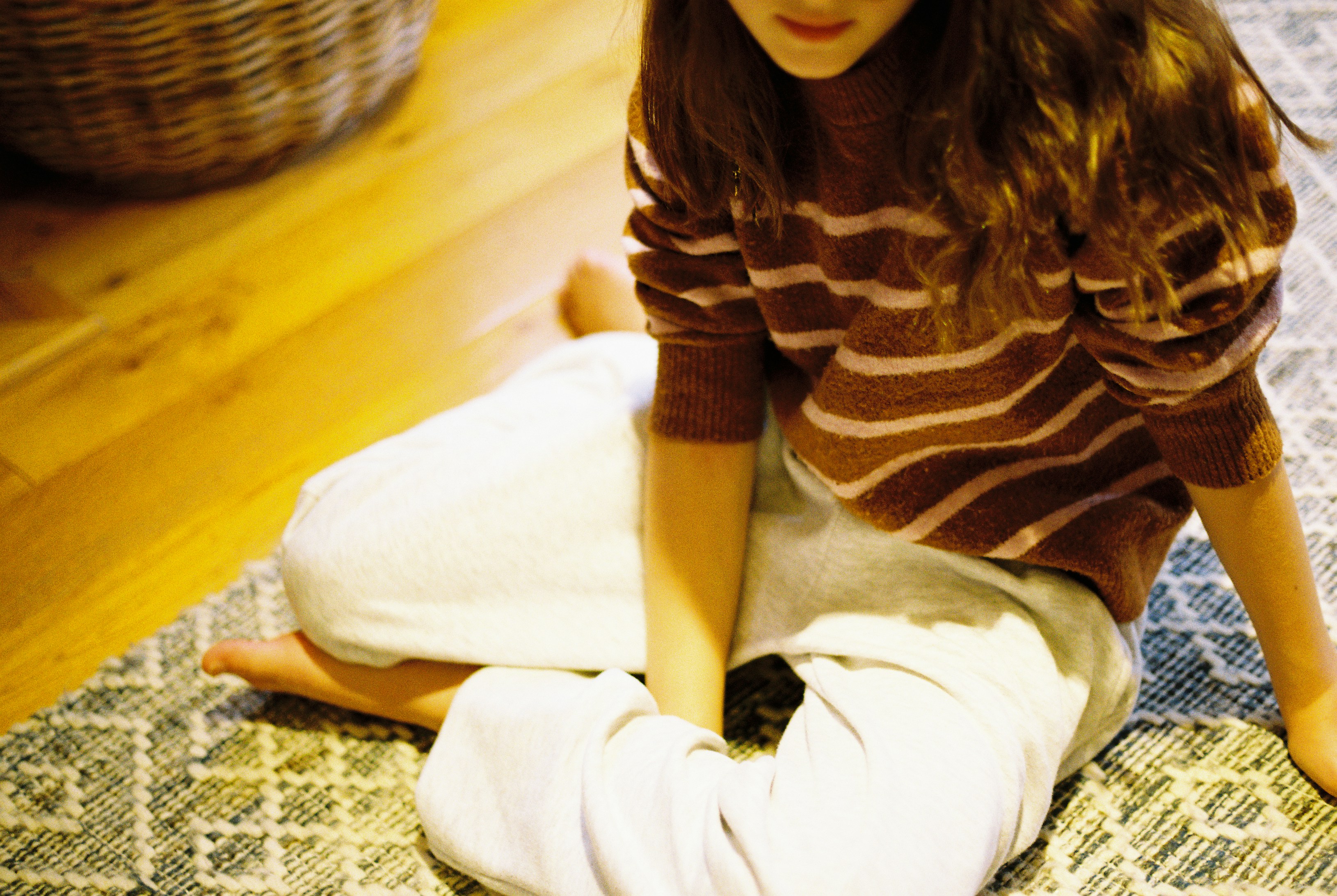 A young girl sits cross-legged on a patterned rug, wearing a striped sweater and light pants, with warm indoor lighting and wooden floor nearby.