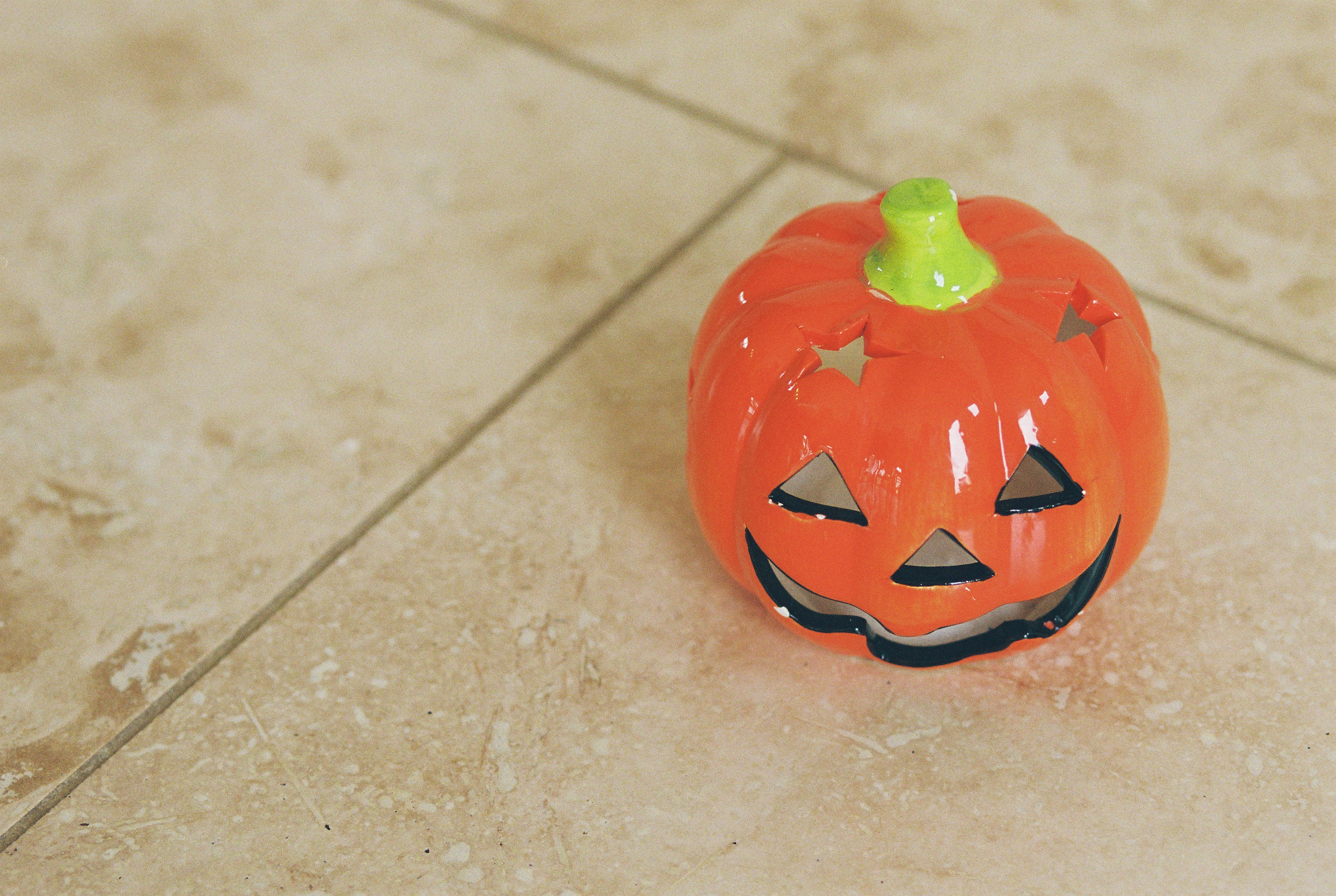 Bright orange jack-o’-lantern resting on light beige tiled floor, small carved face glowing softly; a playful Halloween prop with a glossy surface and a green stem.
