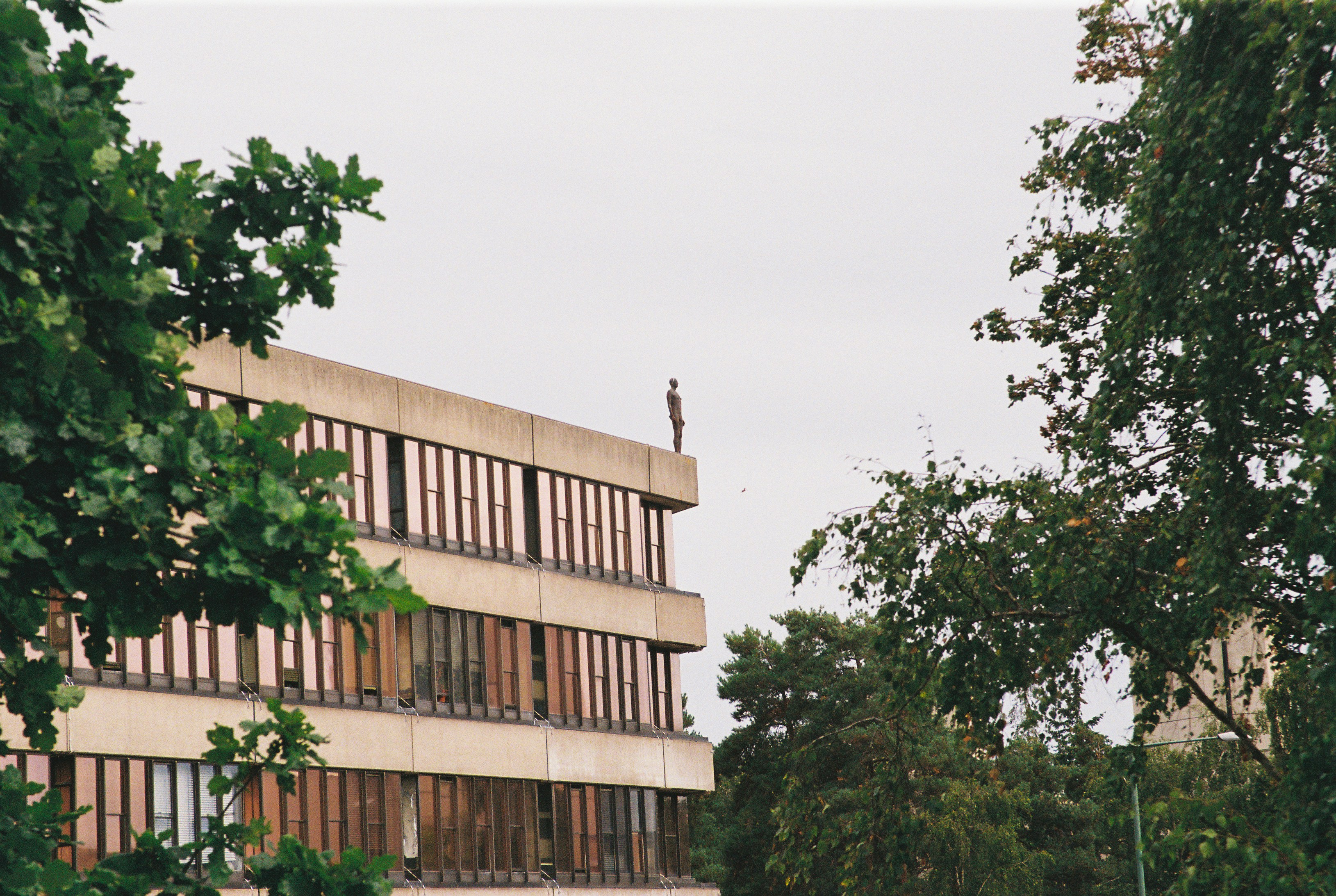 A lone bronze figure sculpture stands atop a brutalist university building, framed by autumn trees.